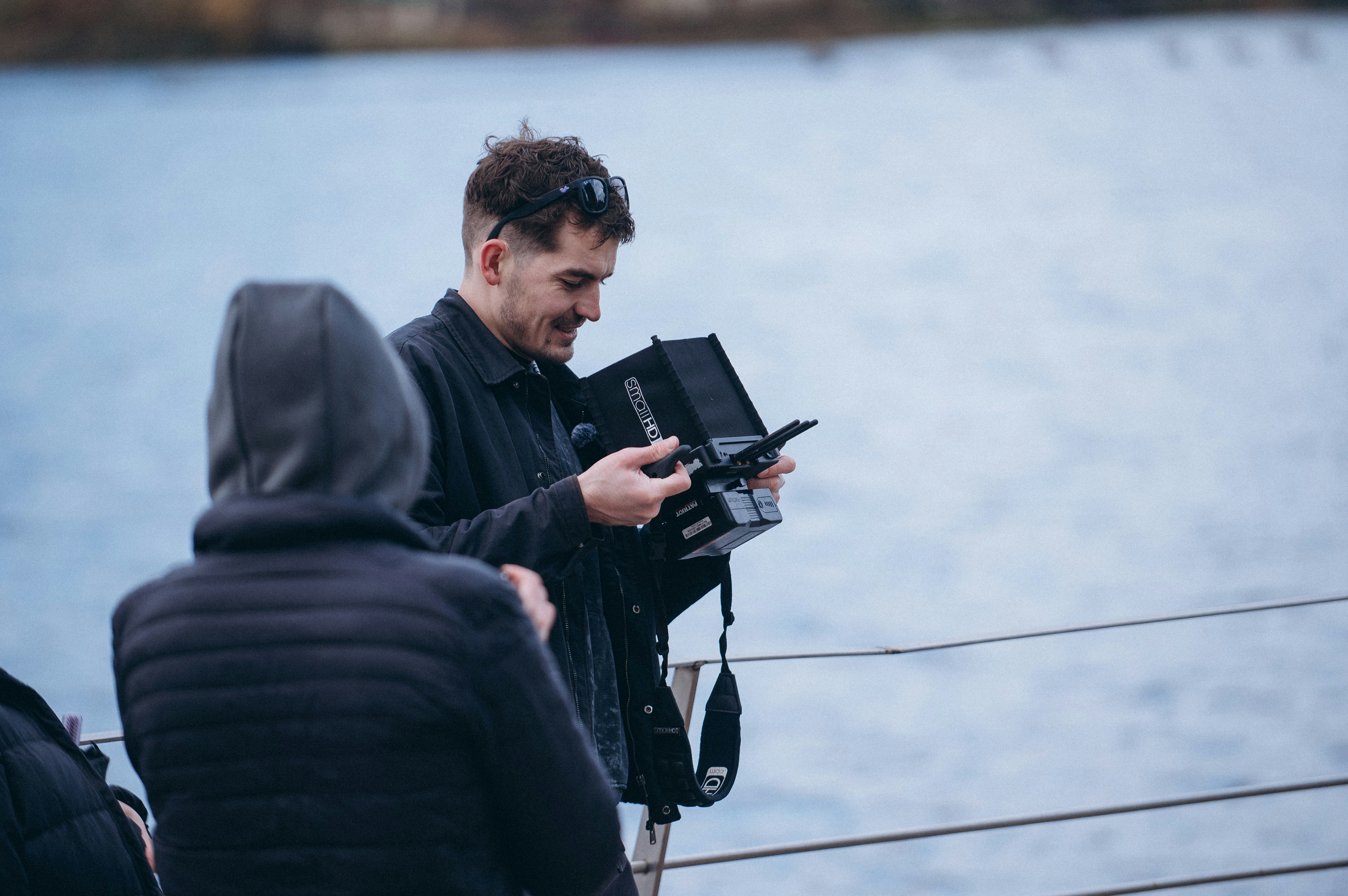A man operating a handheld monitor on a dock by calm water, with another person in a puffer jacket in the foreground; a cable and railing run along the scene, indicating a filming or monitoring setup. This is a candid, real-world shoot.