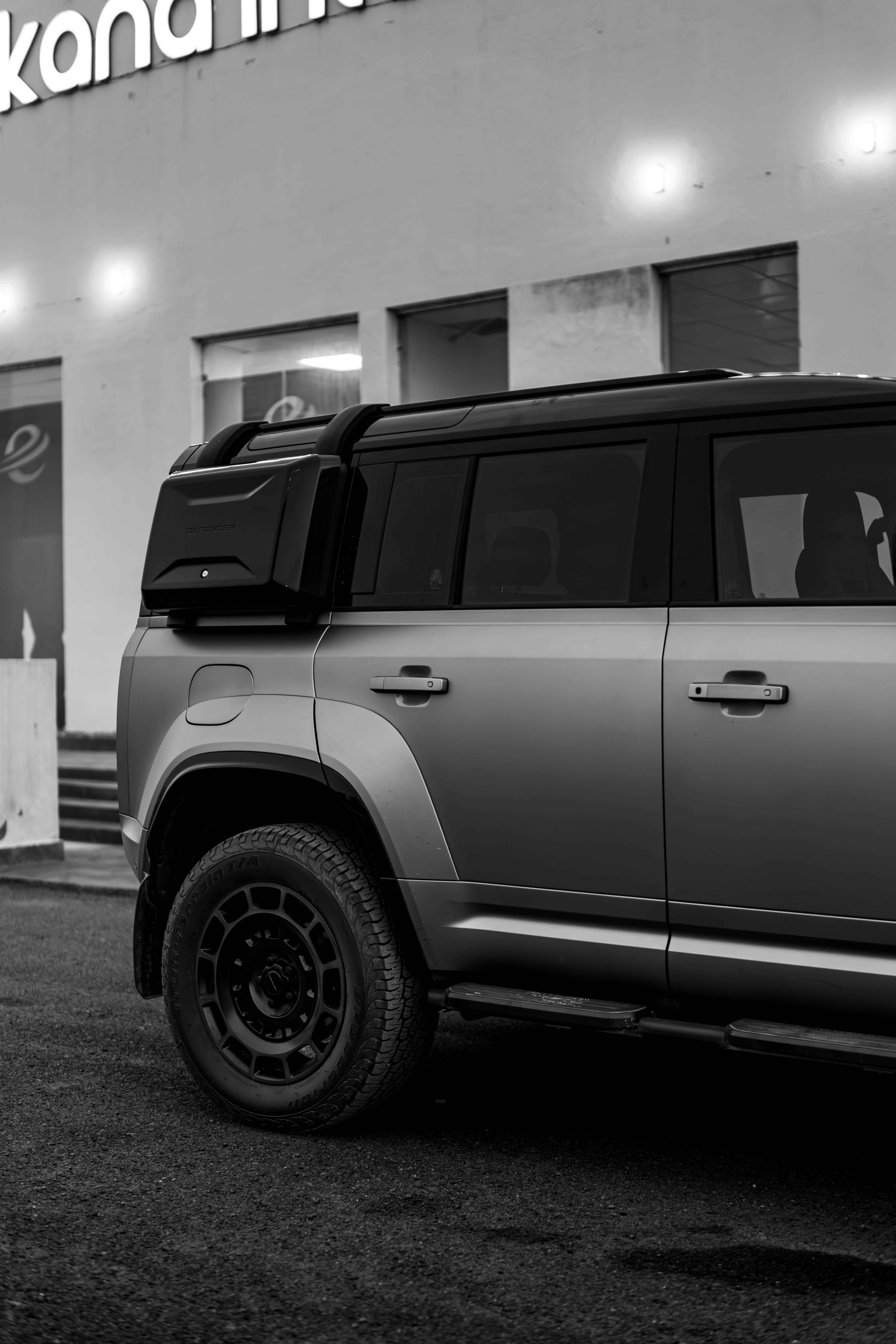 A grayscale side view of a rugged SUV parked beside a building, highlighting the vehicle’s wheel, door lines, and roof details as the scene recedes into the storefronts.