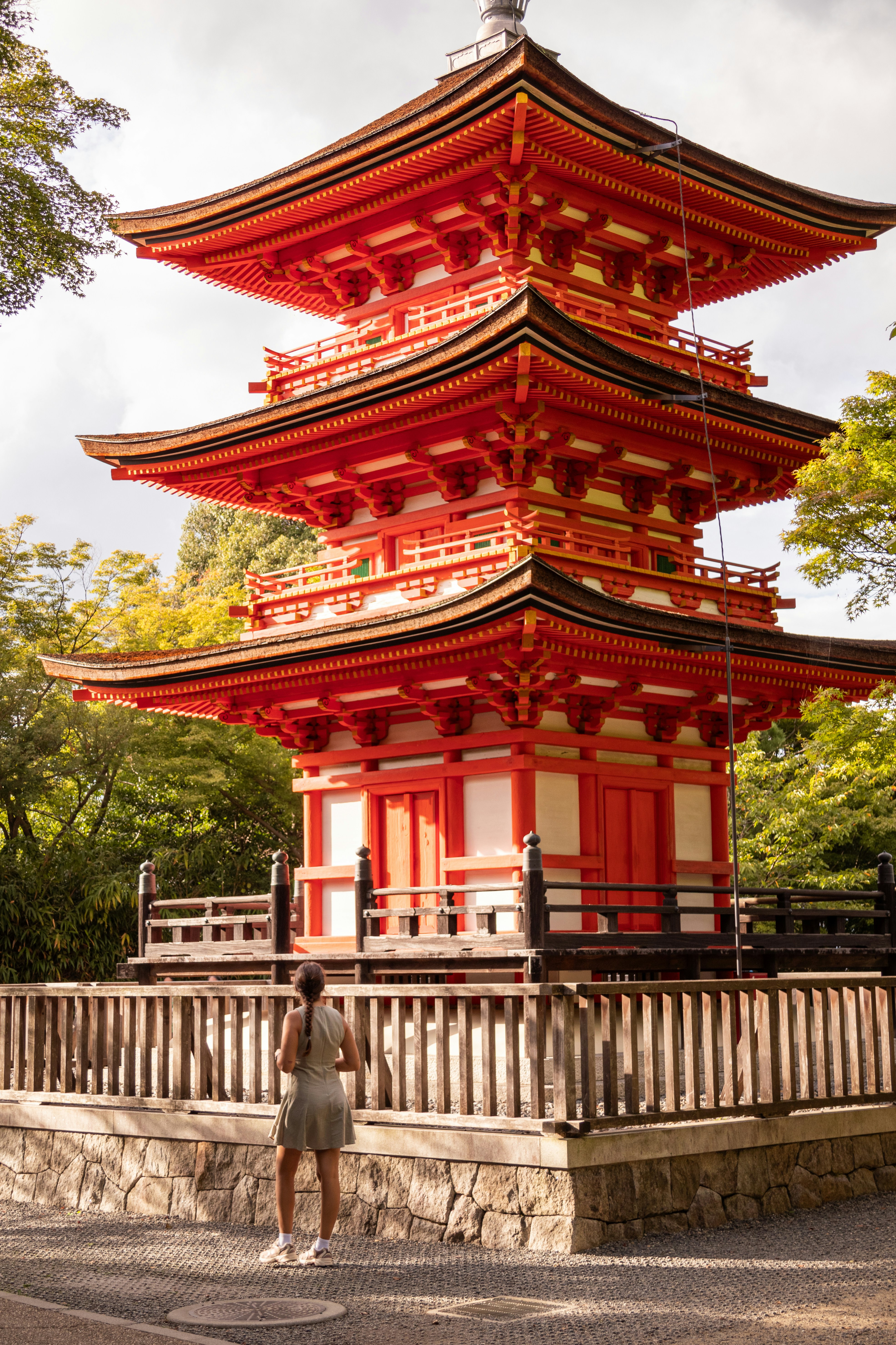 A tall red pagoda rises behind a wooden railing as a lone visitor stands in the foreground, captured in warm daylight; a tranquil temple scene with distinct architectural detailing.