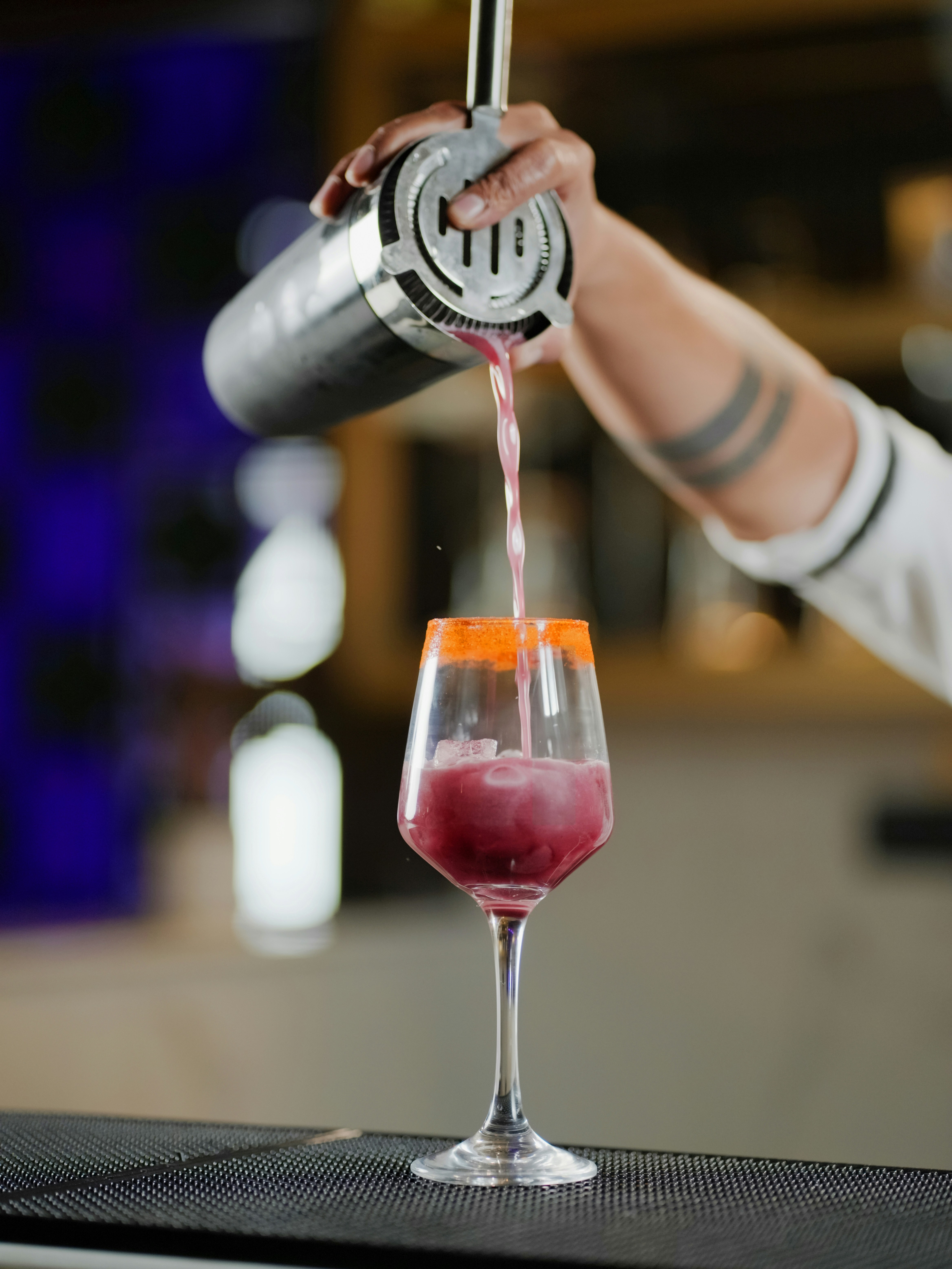 A bartender pours a pink beverage from a sleek nozzle into a stemmed glass on a dim bar counter; the liquid forms a vibrant swirl as the drink settles. The scene emphasizes the craft of a cocktail in a modern bar setting.