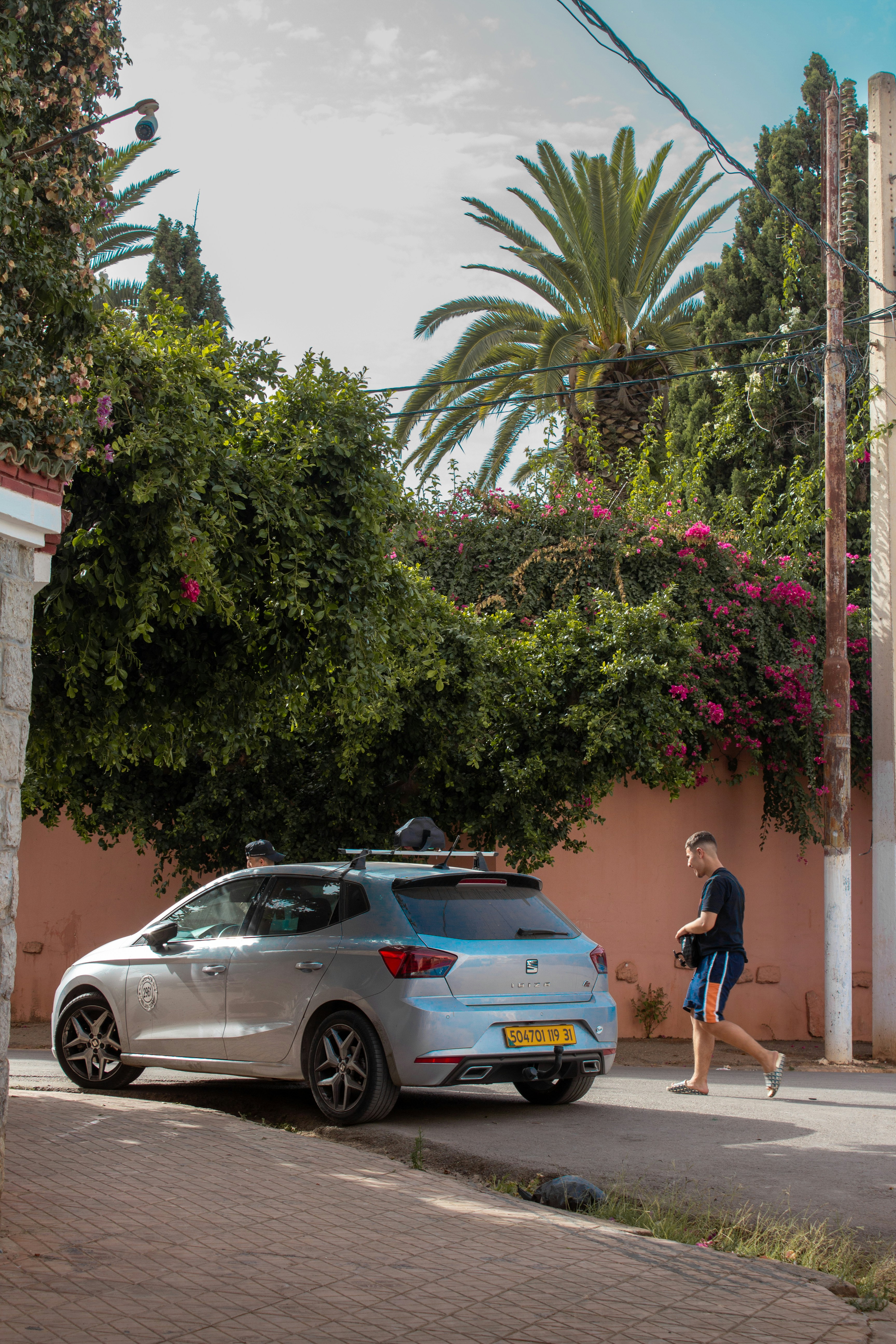 Sunlit Street Scene: Silver Hatchback and Jogger Beneath Blooming Vines