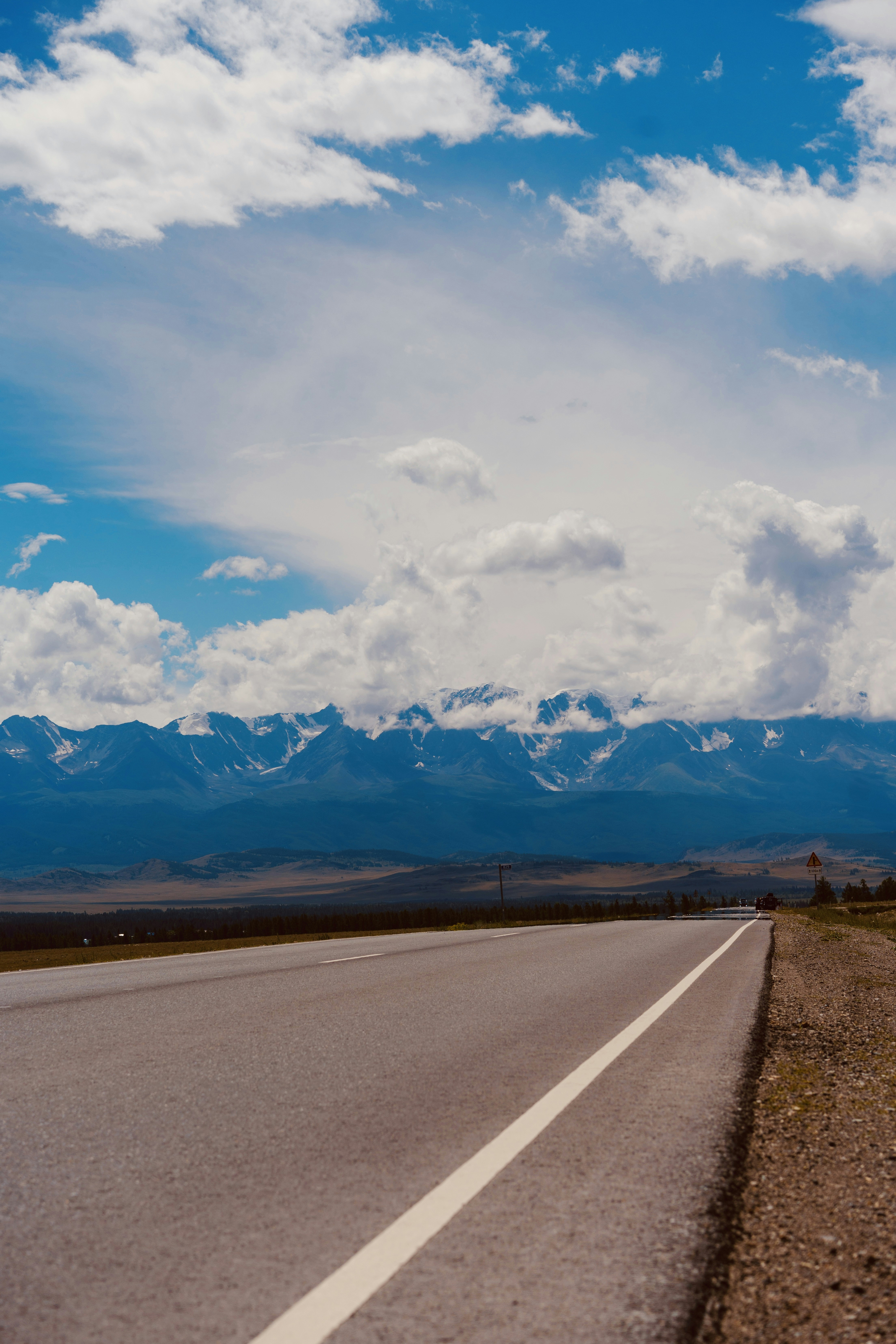 Long straight road converging toward distant mountains under a bright blue sky with scattered clouds. The scene emphasizes vast openness and a sense of journey; a landscape photograph capturing road leading to the horizon.