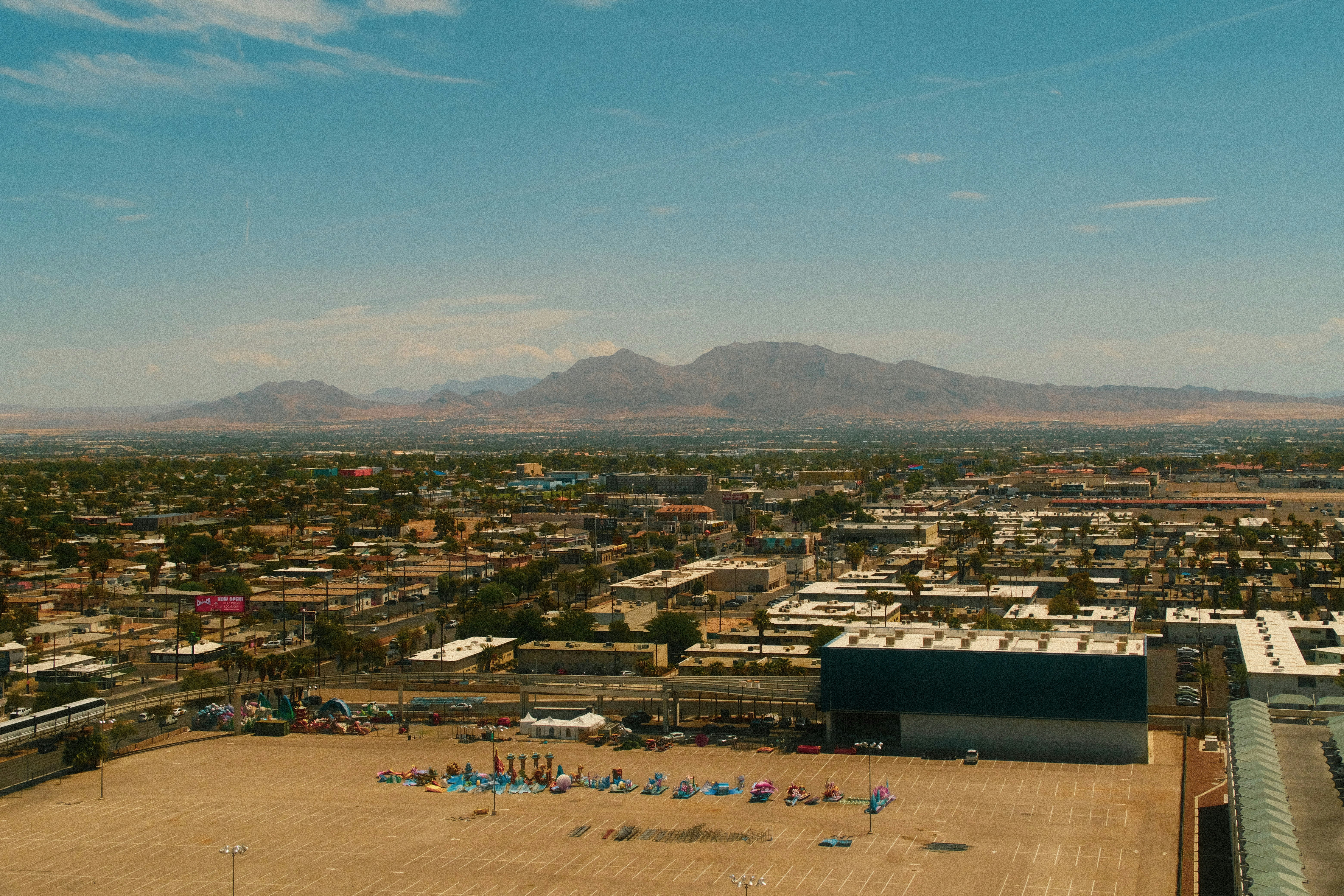Urban landscape with a distant mountain range under a clear sky; foreground shows a large parking/open area with scattered blue umbrellas and a mid-ground row of low buildings.