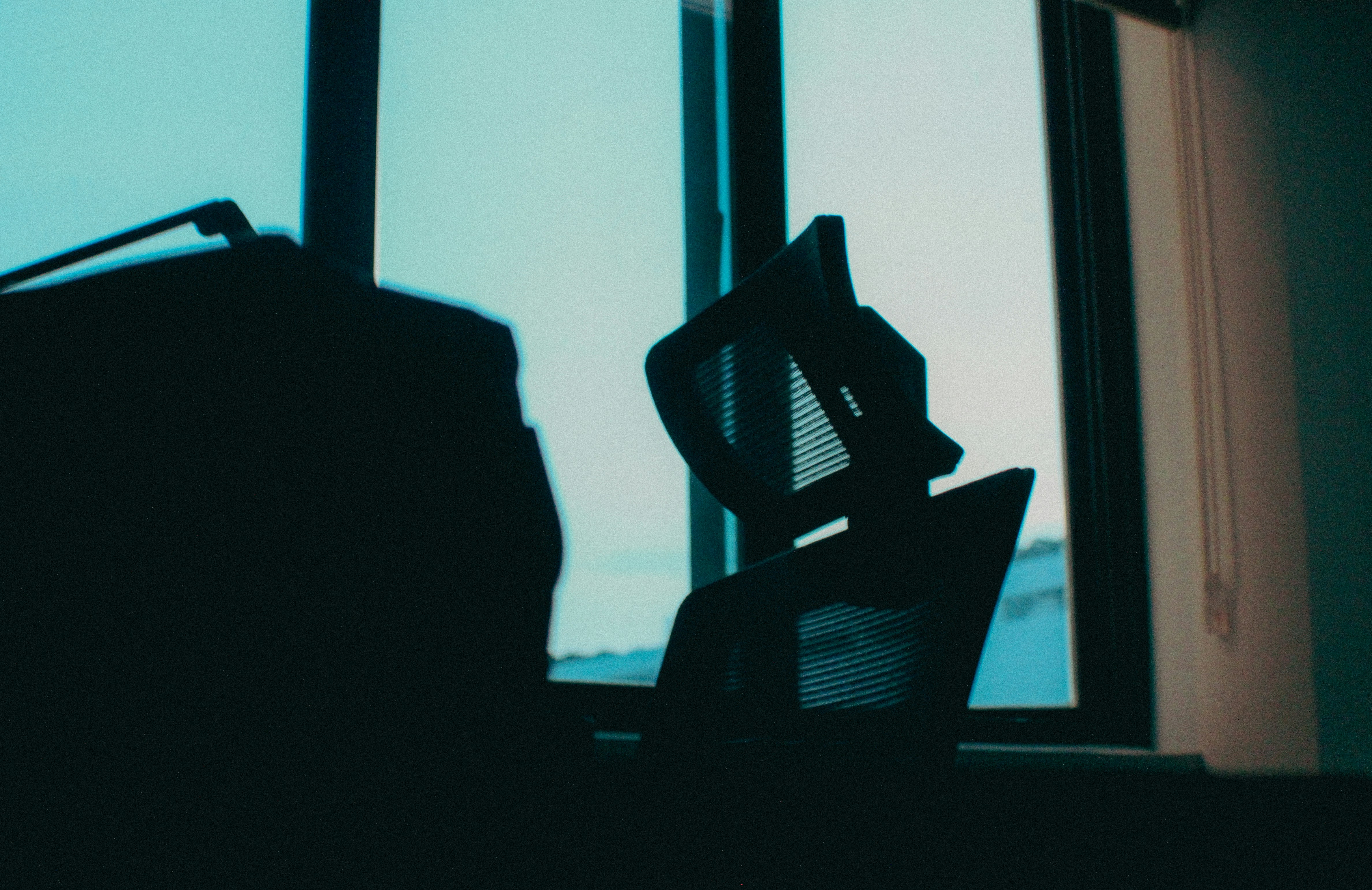Dark silhouettes of an office chair and desk gear sit against a bright window, creating a stark contrast and a moody workspace scene.