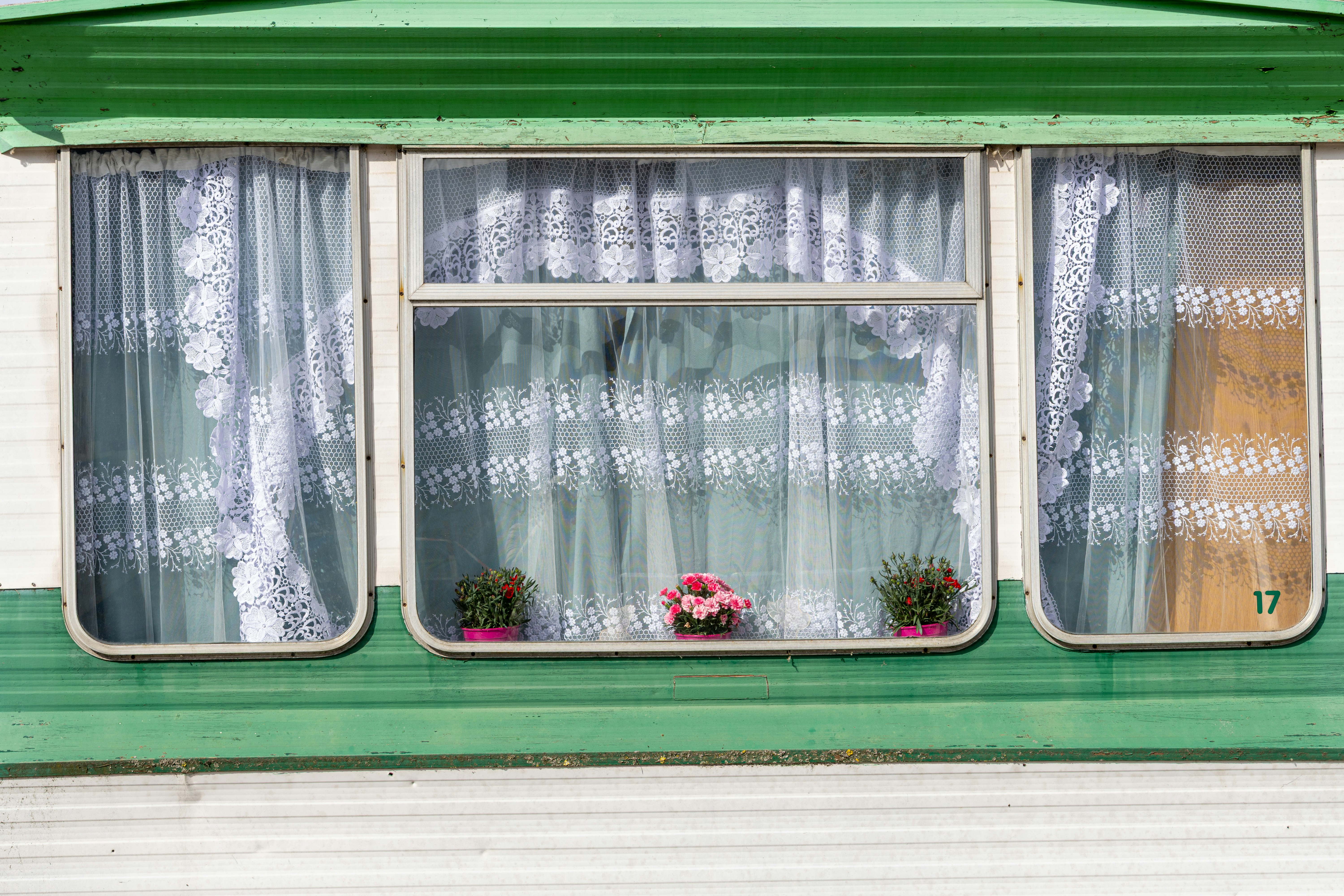 Bright green house façade window with lacy white curtains, small potted flowers on the sill, and a numbered pane hinting at a living space.