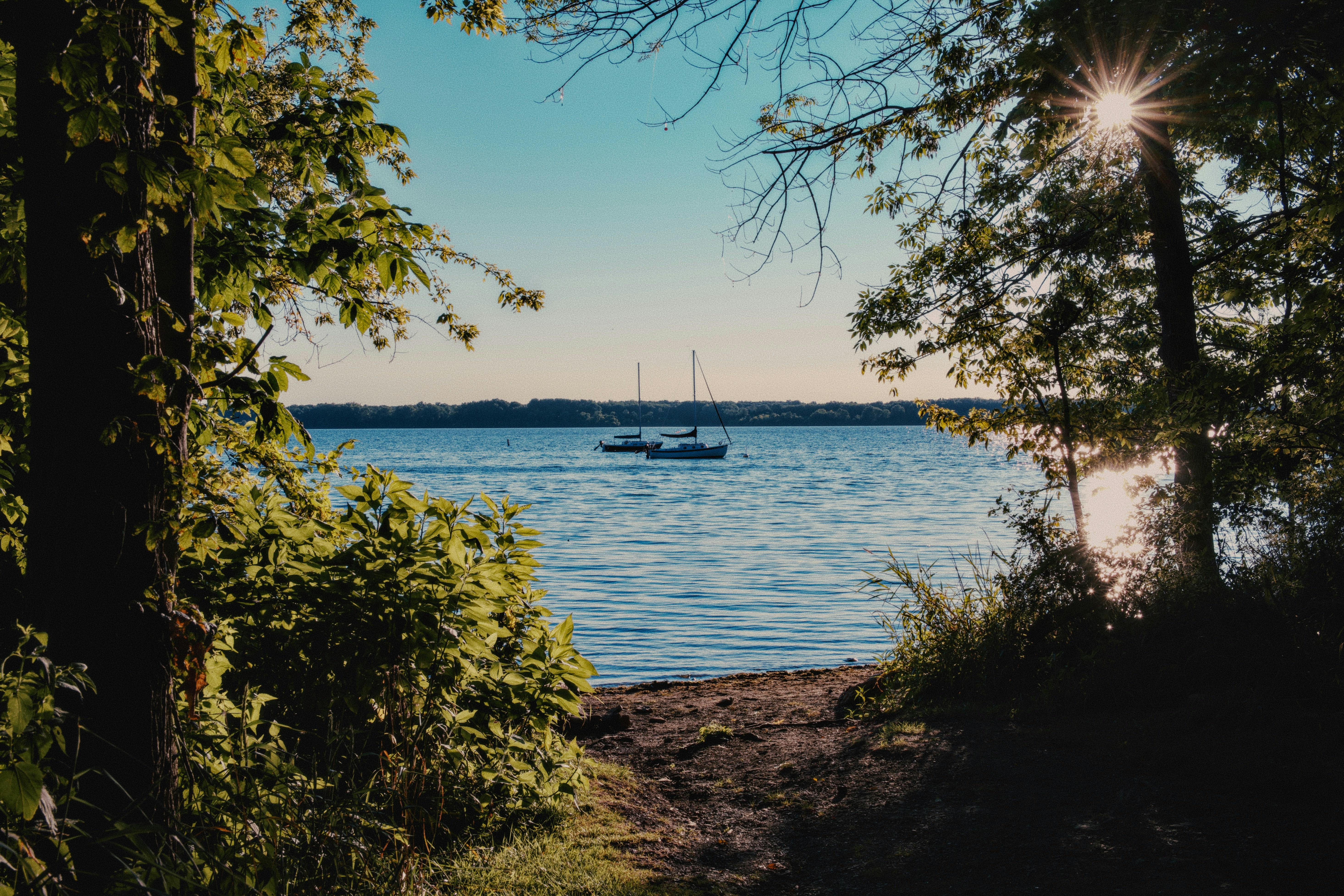 Sunlit boat drifting on calm lake framed by leafy shores and a bright sky. A tranquil landscape with edge detail of trees and a sunburst reflection on the water.