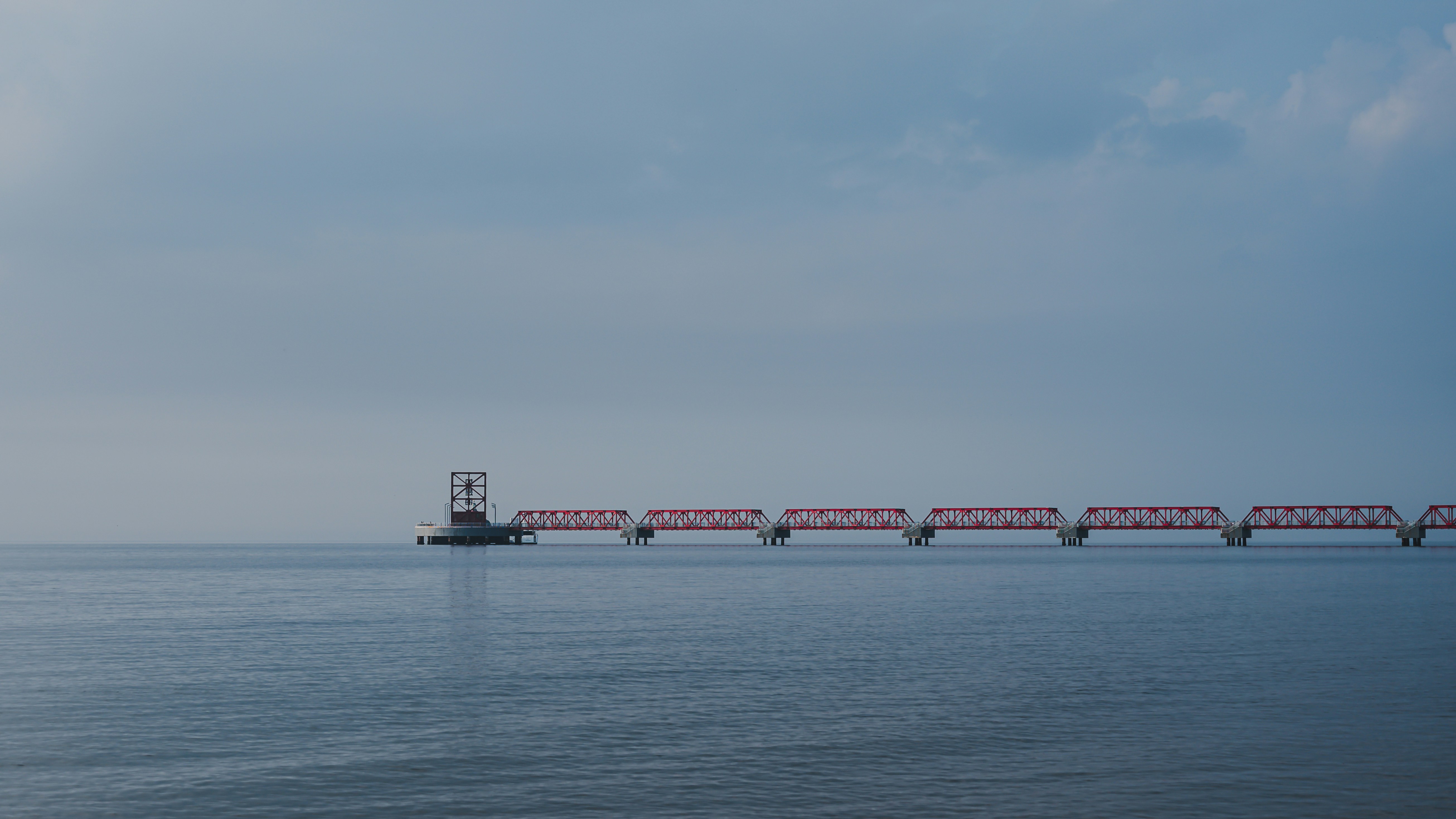 Distant freight train cars traverse a flat horizon over calm sea, under a soft, clouded sky. A minimal seascape emphasizes line and negative space with a hint of structure on the water.