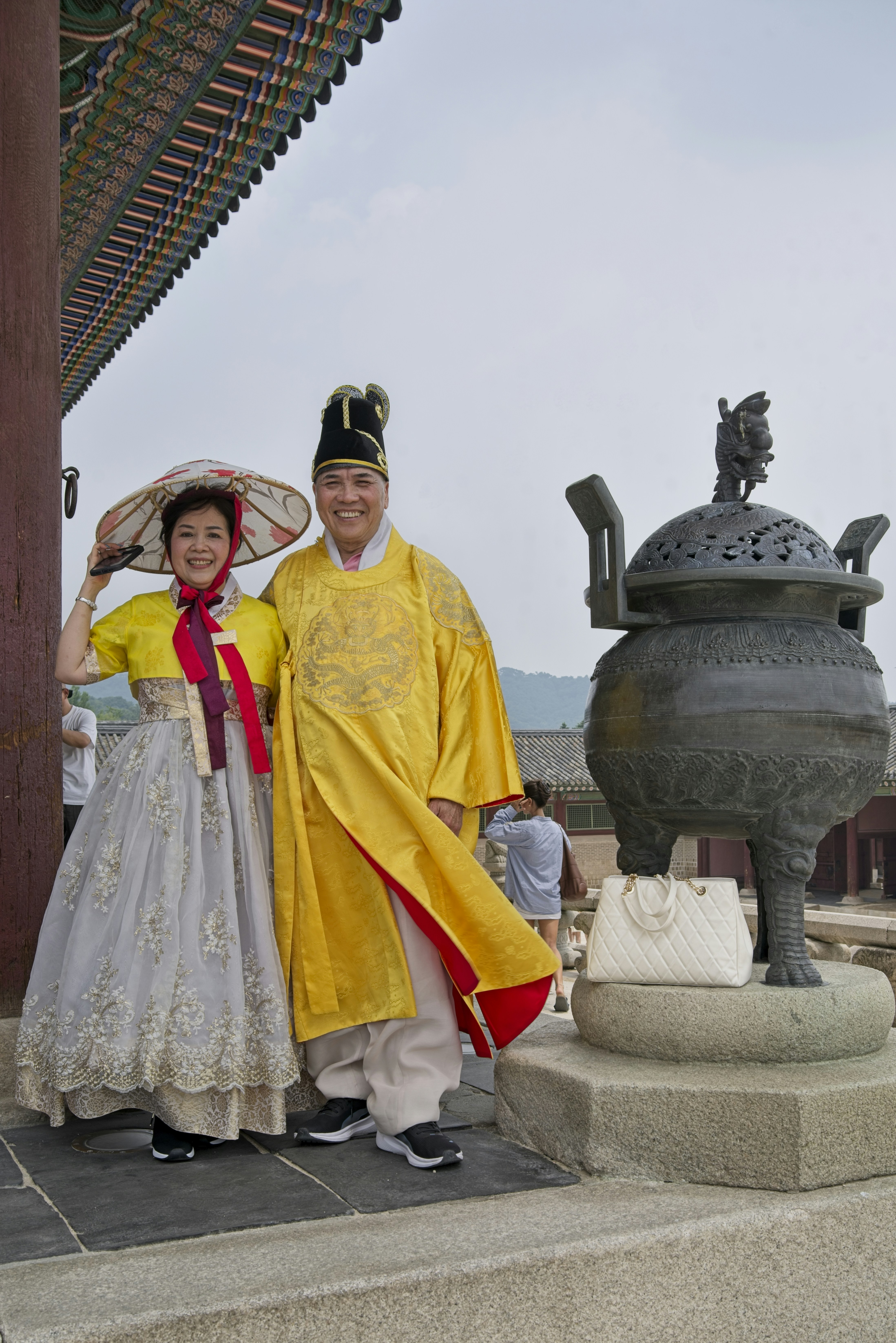 A smiling couple pose beneath a painted palace eave, dressed in elaborate hanbok — the man in a golden dragon-embroidered robe and black official's hat, the woman in an elegant gray gown topped with a wide woven hat. Beside them, a massive bronze tripod incense burner with an ornate dragon finial stands on its stone pedestal, a modern white handbag incongruously perched at its base. Distant palace rooftops and hazy mountains complete the scene, blending playful costume tourism with Gyeongbokgung's timeless architectural grandeur on a warm July afternoon.