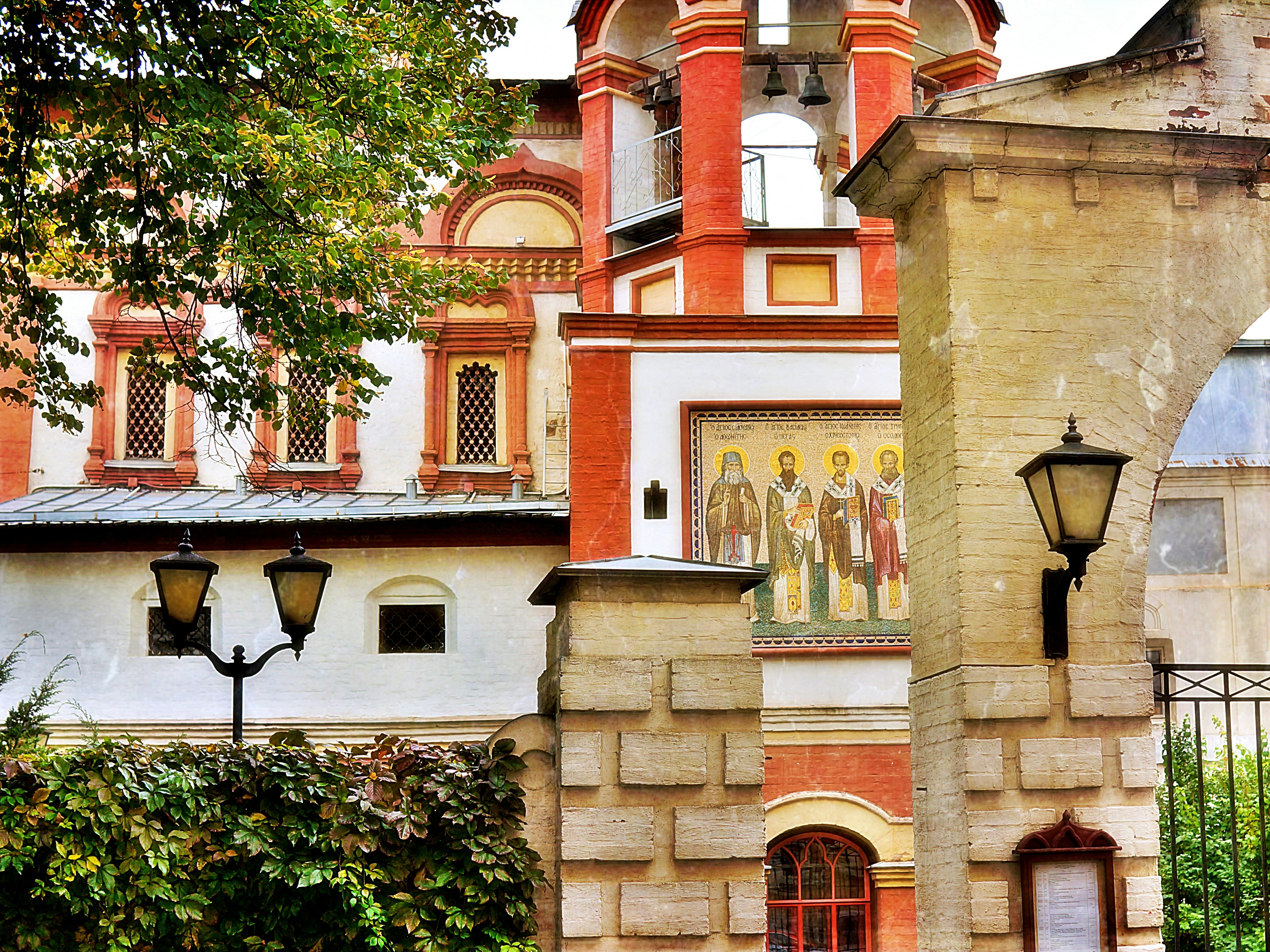 Colorful church facade with red brick, arches and a mosaic of saints, framed by a stone arch and greenery.