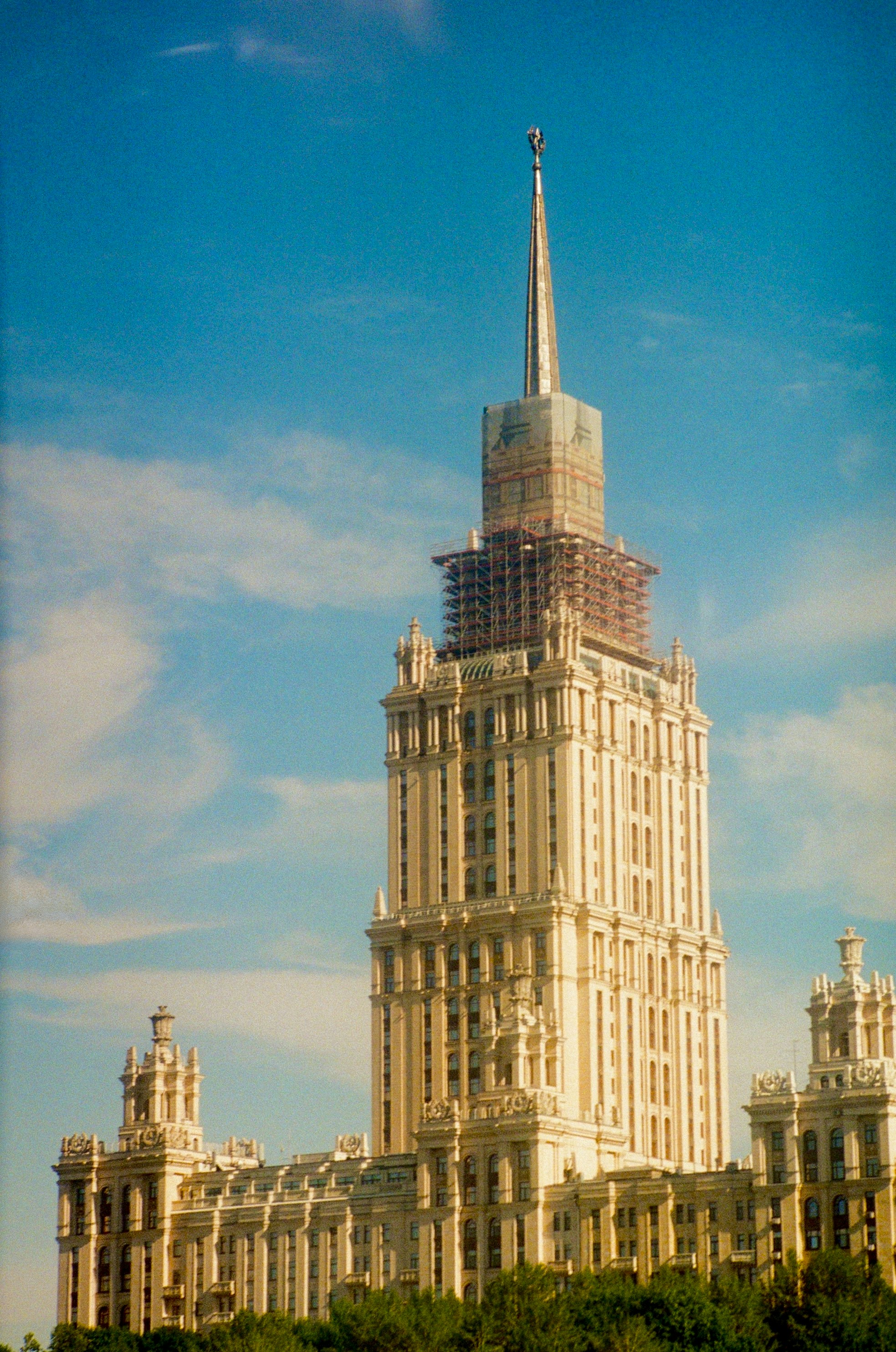 Tall historic skyscraper with a spire under construction against a blue sky; foreground shows greenery and lower wings revealing detailed architectural ornamentation.