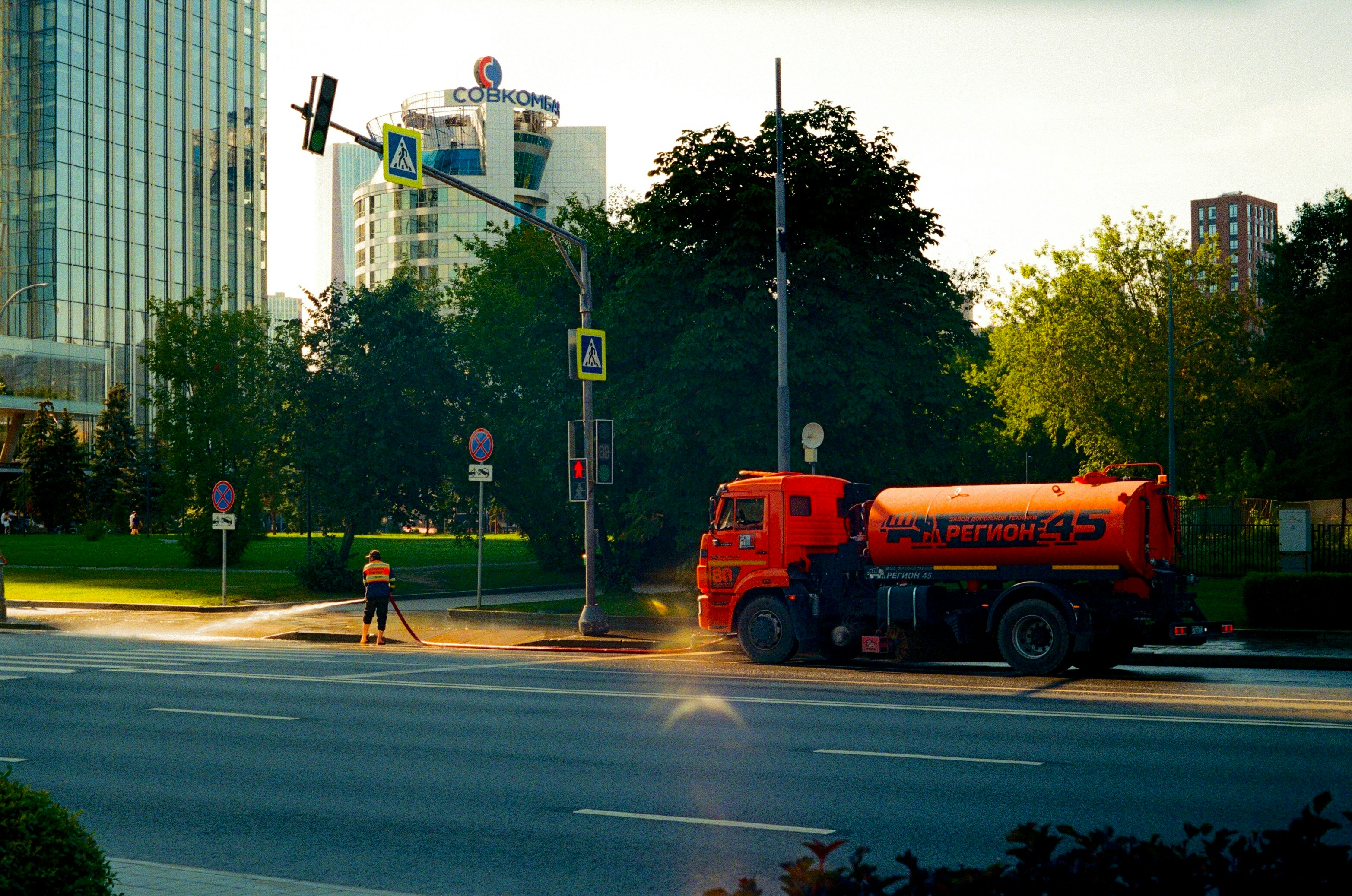 Red tanker truck parked at a crosswalk in a sunlit city street, with a pedestrian and modern buildings in the background.