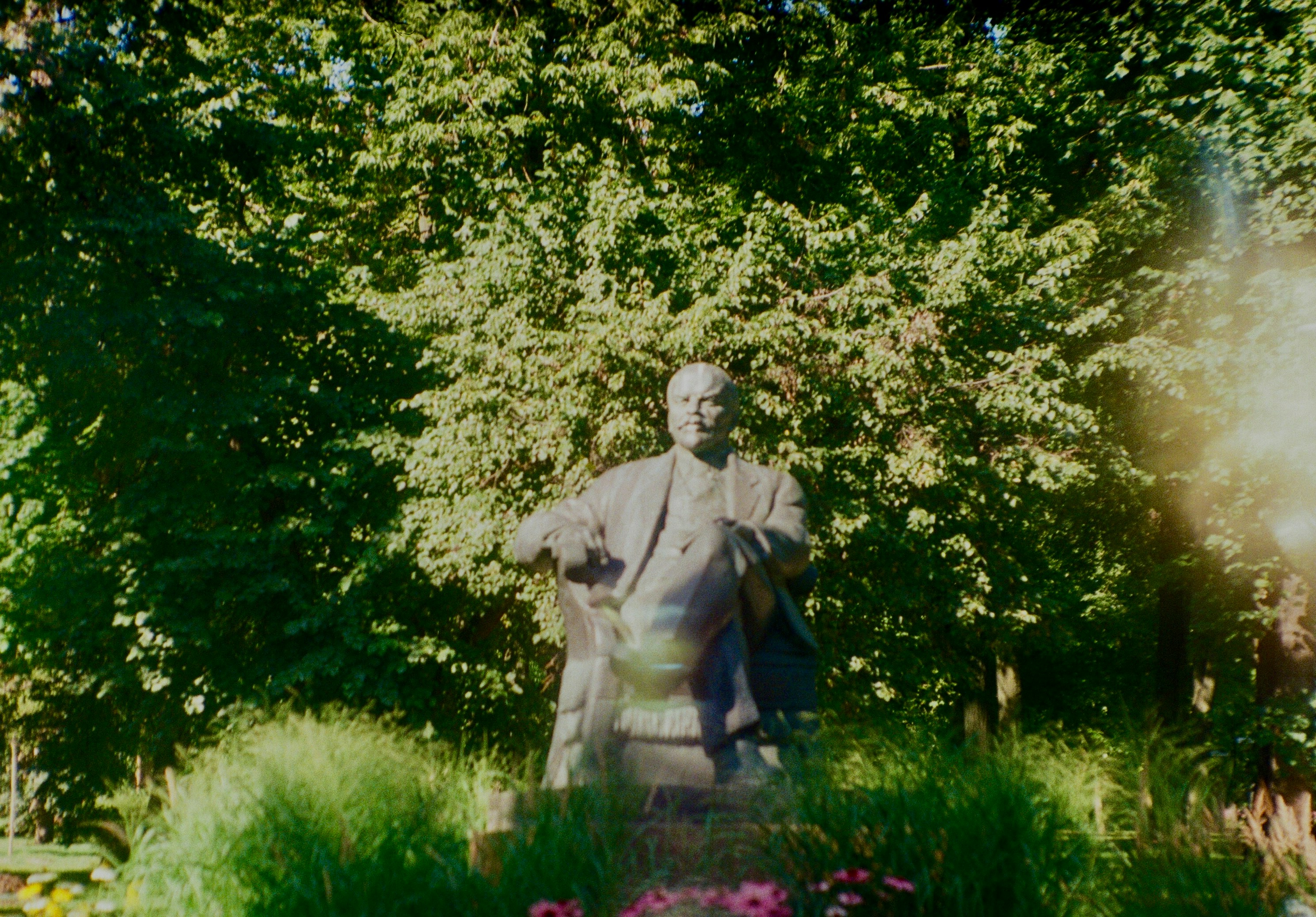 Stone statue surrounded by lush garden in bright daylight; a monument framed by trees and soft sunlight filtering through leaves.