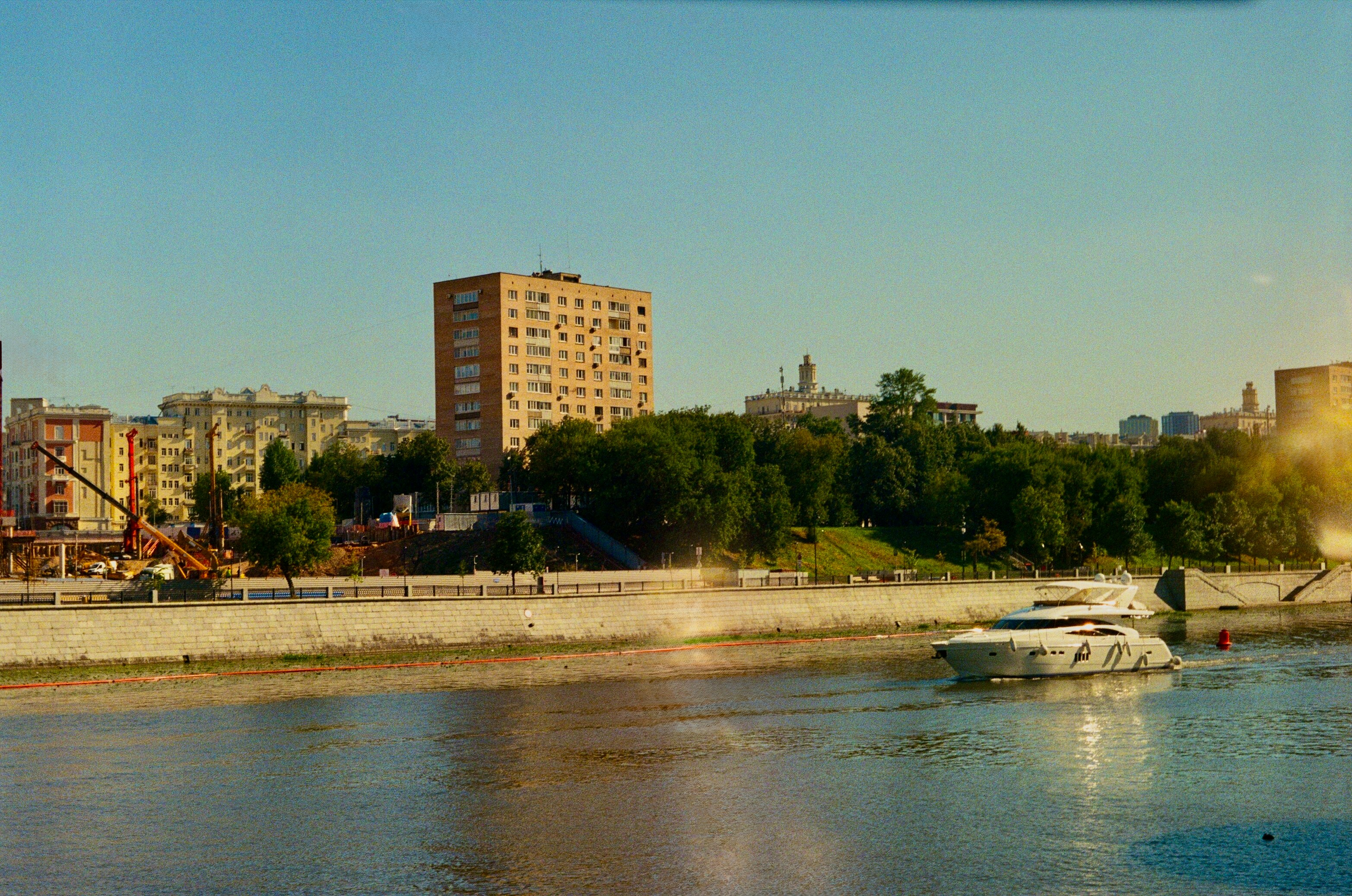 City riverside with a grassy promenade and a white motor yacht on calm water; a mix of mid-rise buildings and trees forms the backdrop on a clear day.