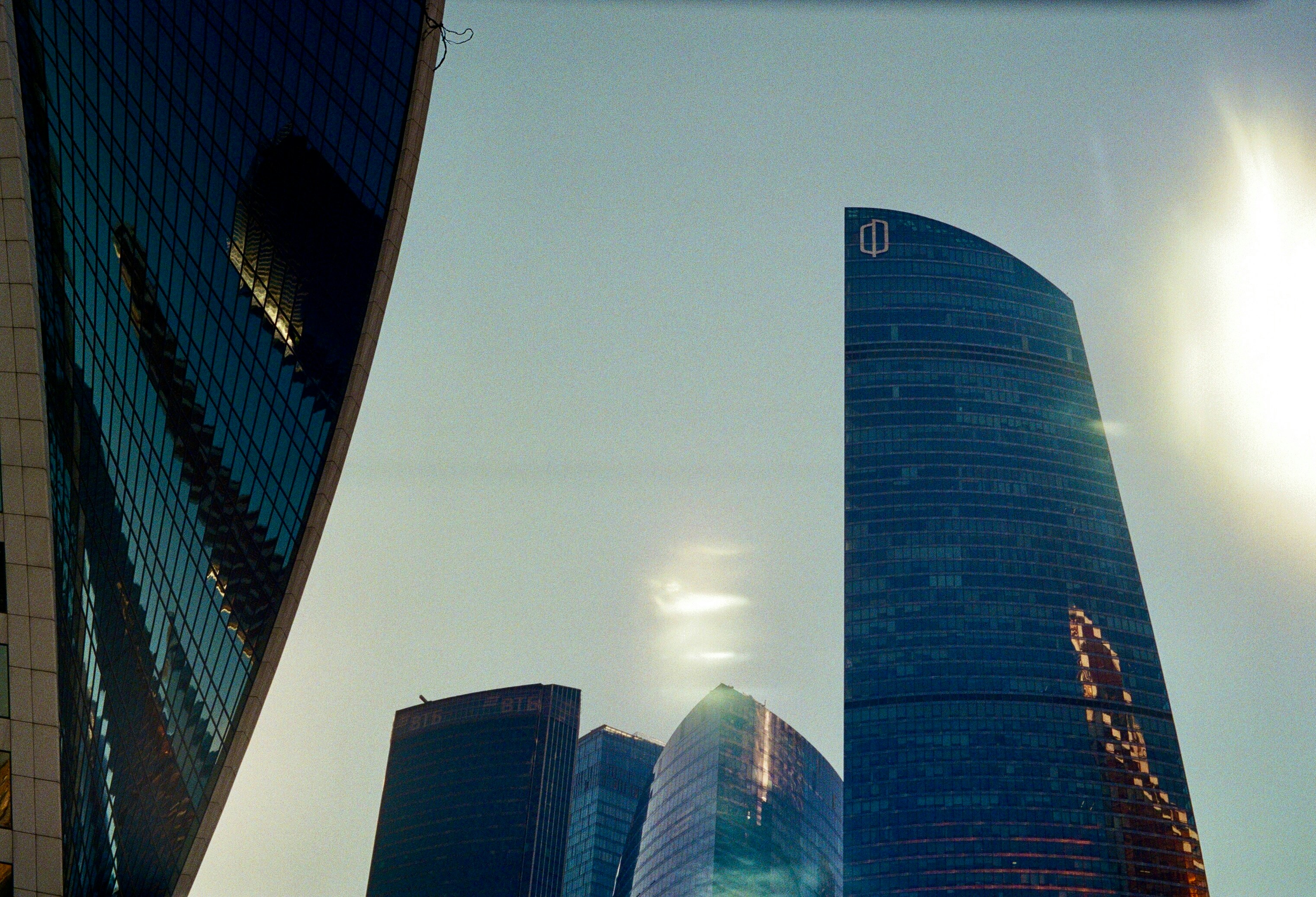 Sleek glass skyscrapers rise against a pale sky, with sunlit reflections visible on curved facades and a hint of surrounding structures.