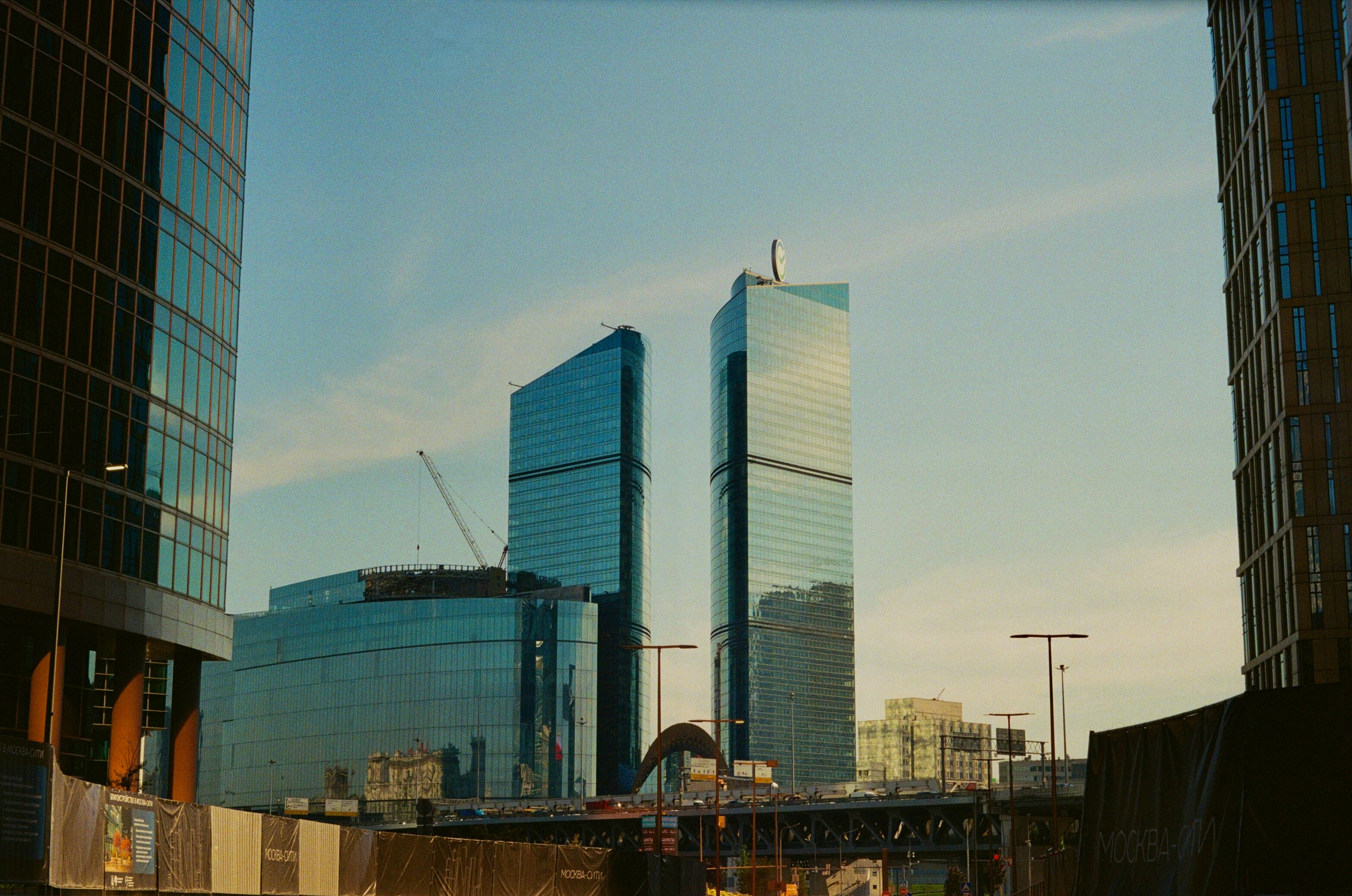 Urban skyline with modern glass towers rising over a low foreground of highway and structures; a bright blue sky underscores the reflective façades.