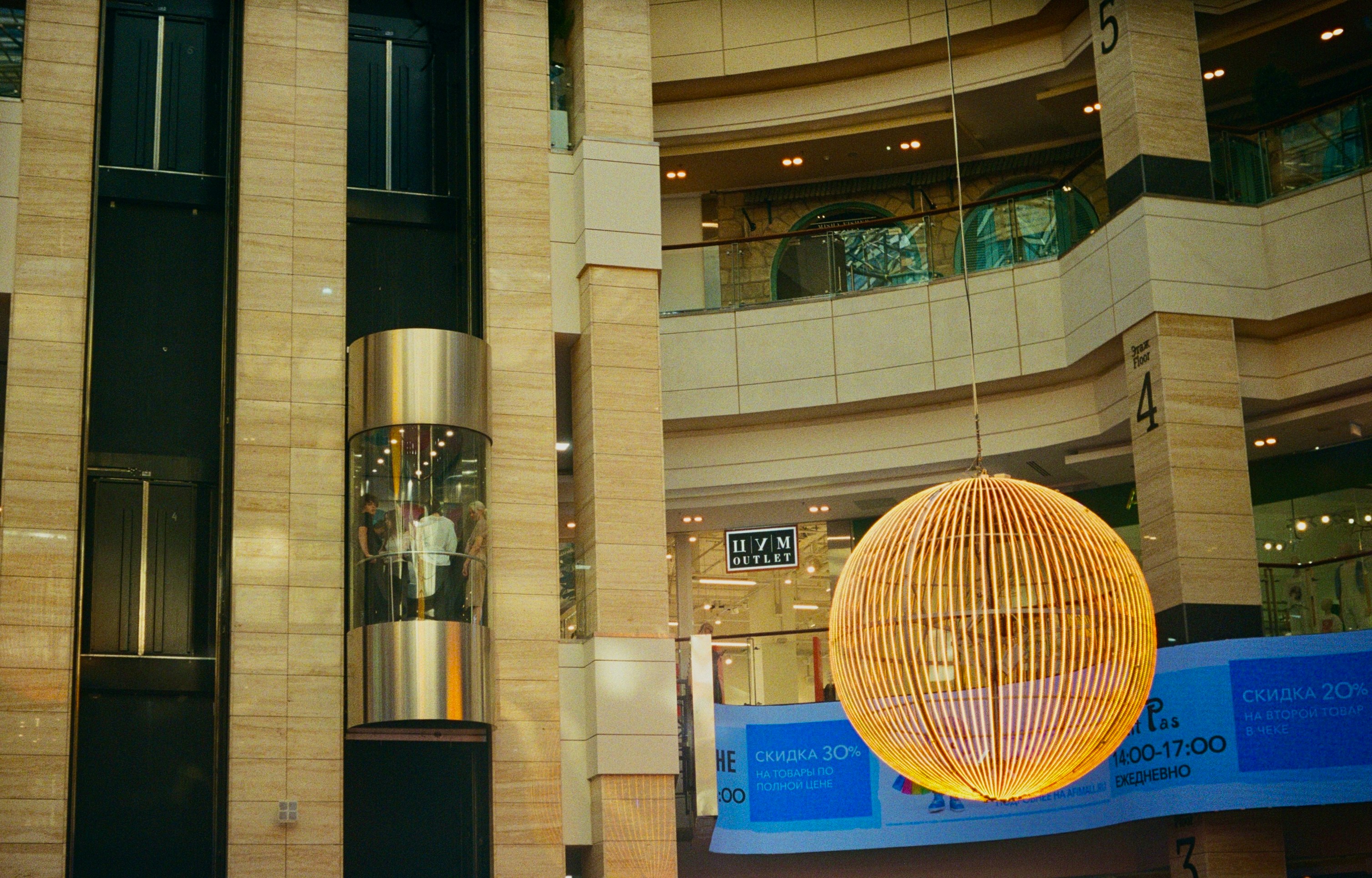 A warm spherical light hangs in a spacious atrium with architectural columns and blue banners in the background. The scene highlights interior design and the sculpture-like lamp as the focal element.