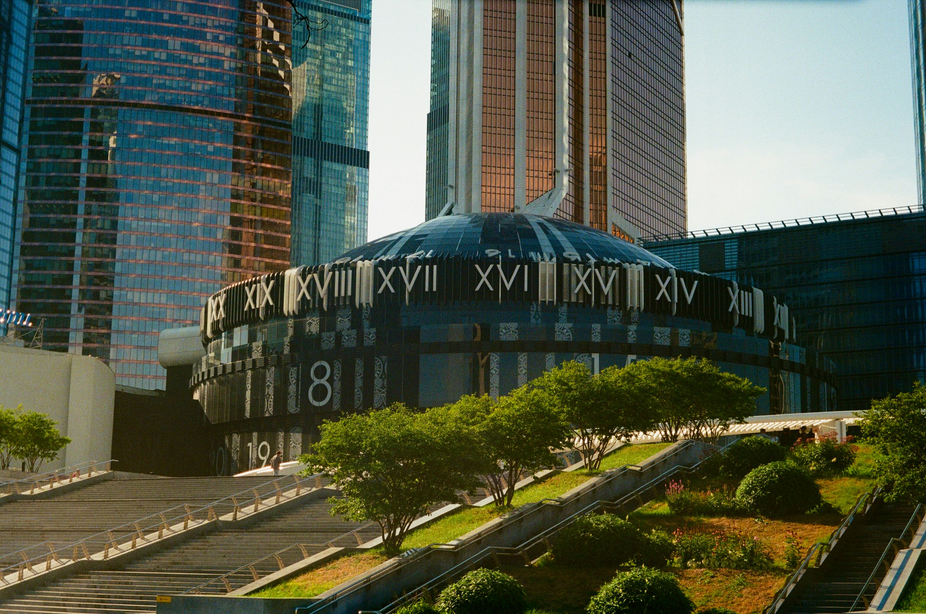 A futuristic circular structure with roman numerals sits amid glass towers and landscaped stairs, suggesting a public landmark or clock-inspired pavilion.