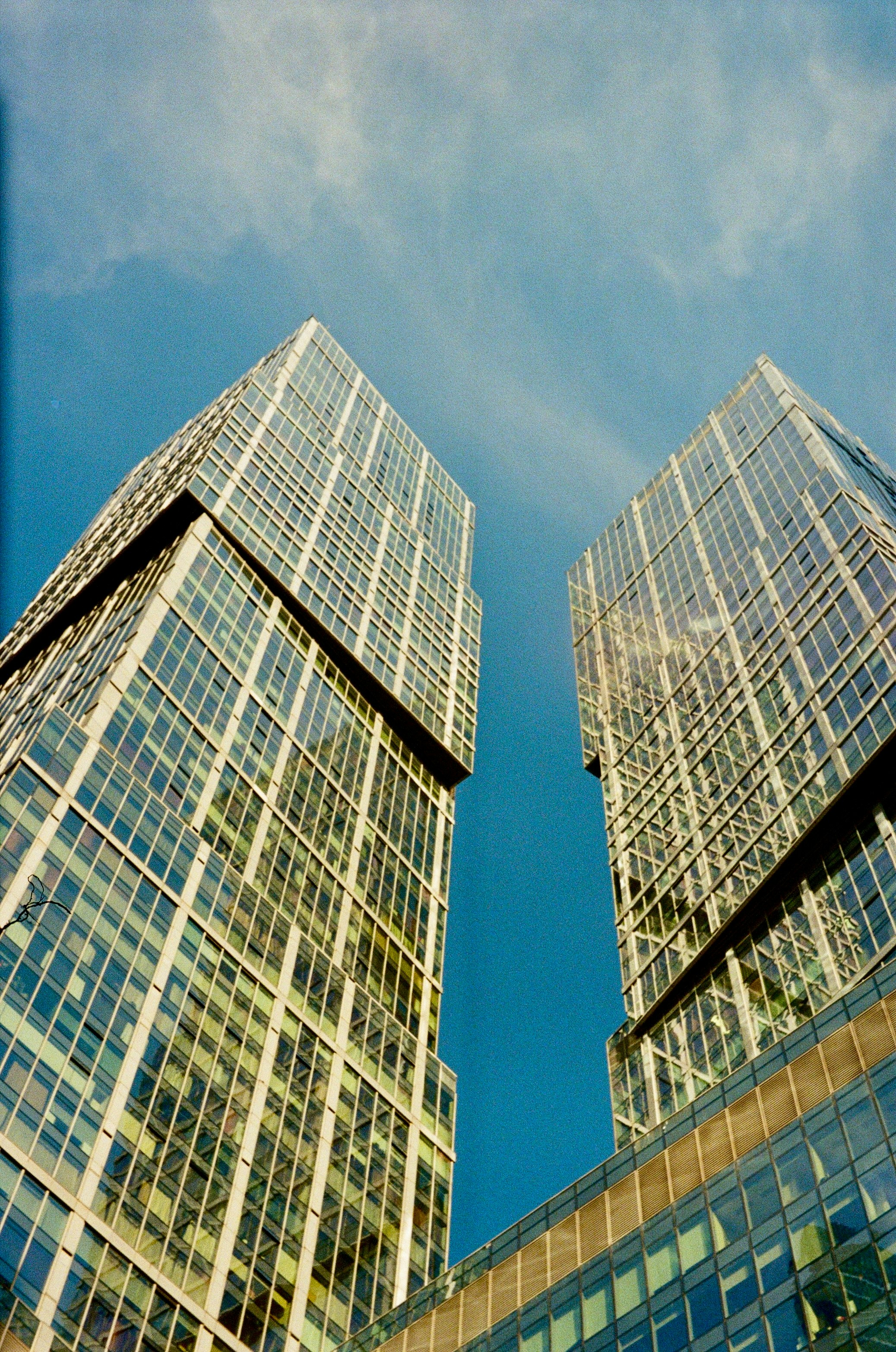 Two glass-clad skyscrapers stretch upward against a bright blue sky, their reflective facades capturing surrounding light and geometry. This architectural shot emphasizes vertical lines and modern glass architecture.