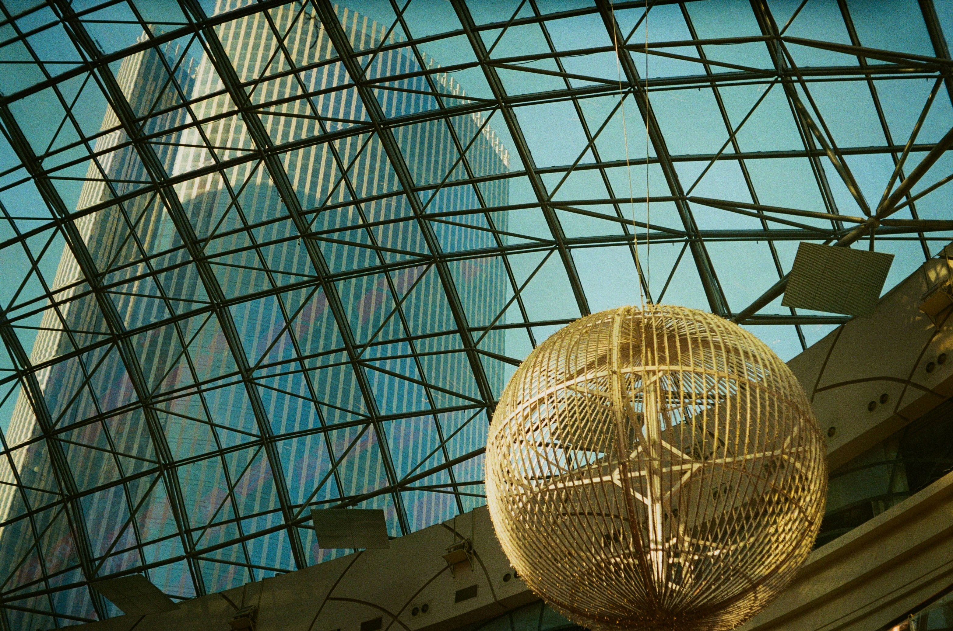 A film photograph taken inside a shopping mall in Moscow City, looking up through a glass roof that frames a towering glass skyscraper above. A decorative hanging sphere suspends in the lower part of the frame, adding depth to this layered, modern architectural scene.