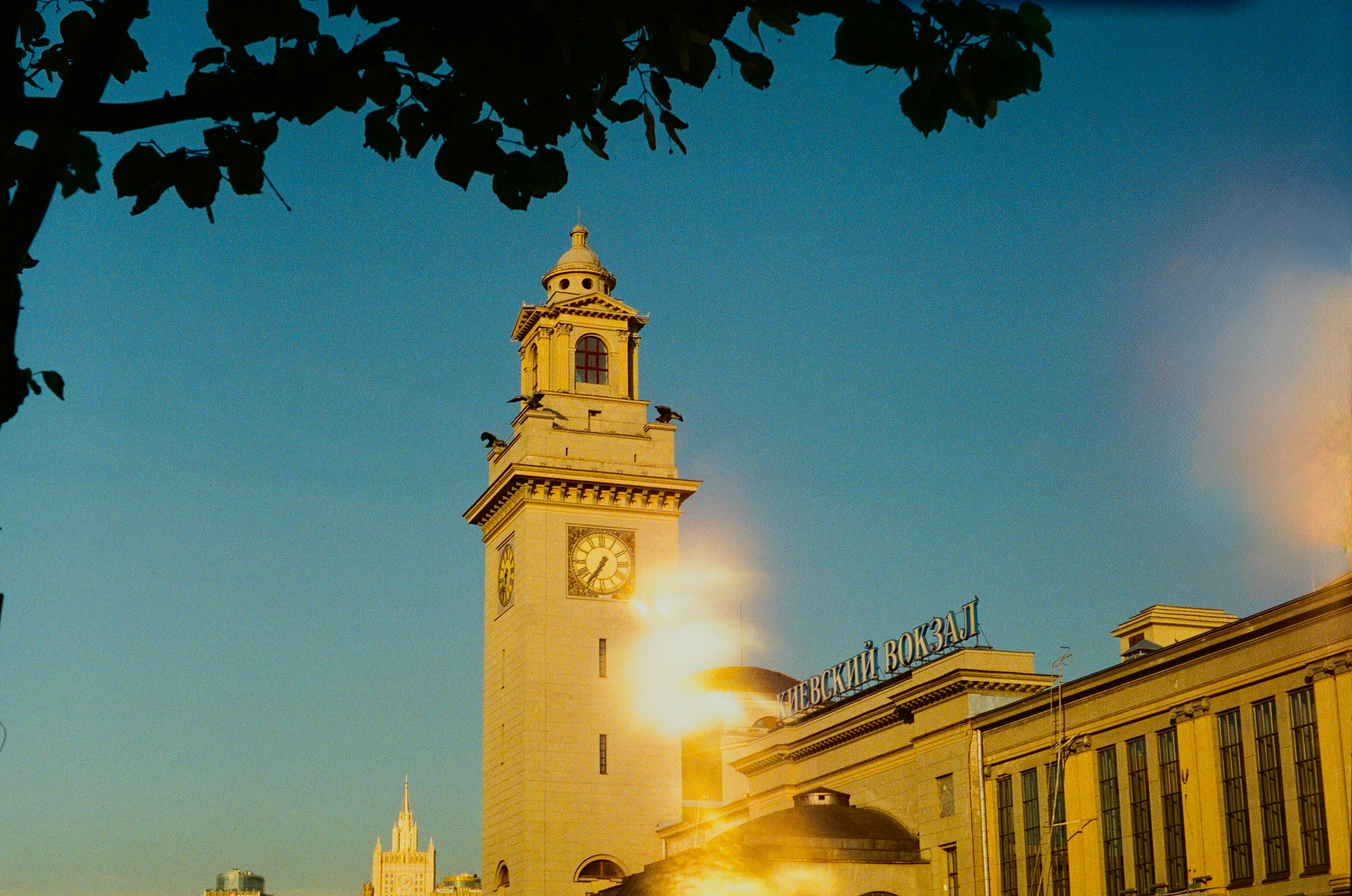 Sunlit clocktower rises over a classic rail station facade with warm glow and a bright sky, capturing urban architecture and motion blur near a passing tram.