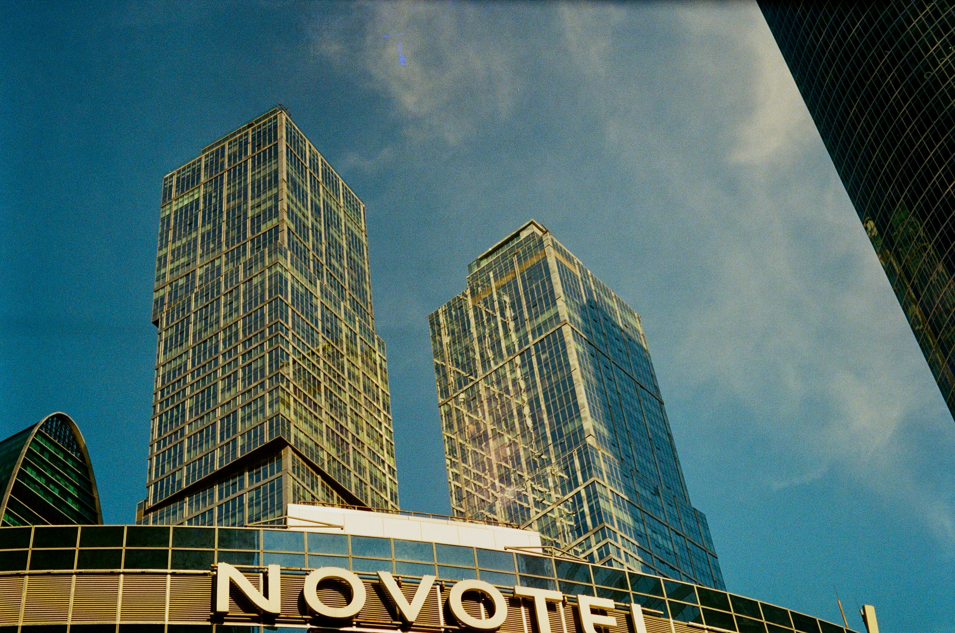 High-rise glass towers rise above a curved hotel facade with a prominent NOVOTEL sign beneath; blue sky with a few clouds forms a crisp urban scene.
