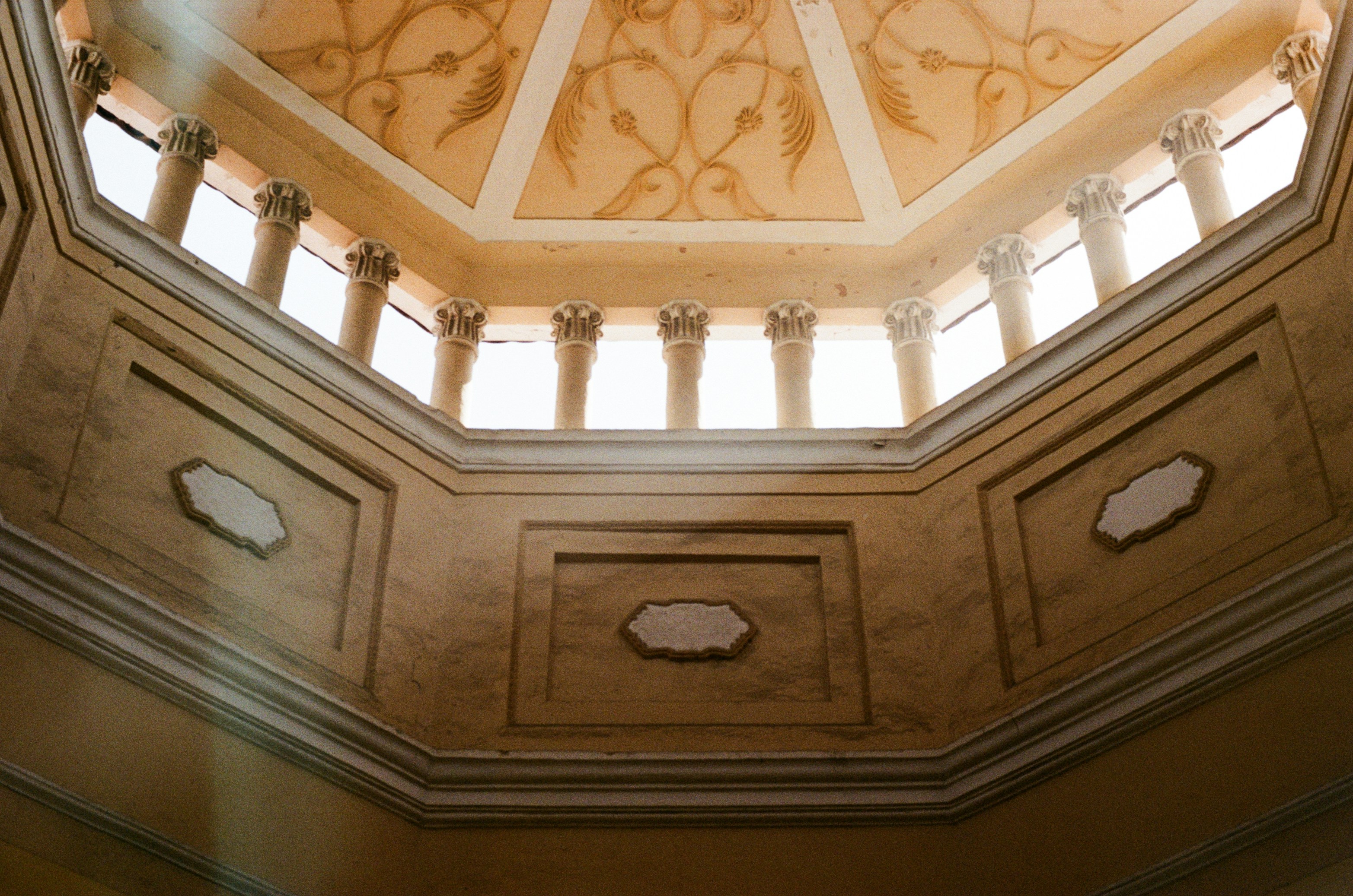 Architectural interior with an ornate domed ceiling lined by columns; warm-toned marble walls and decorative coffers. A view upward toward the skylight arcade, capturing the symmetry and classical detailing.