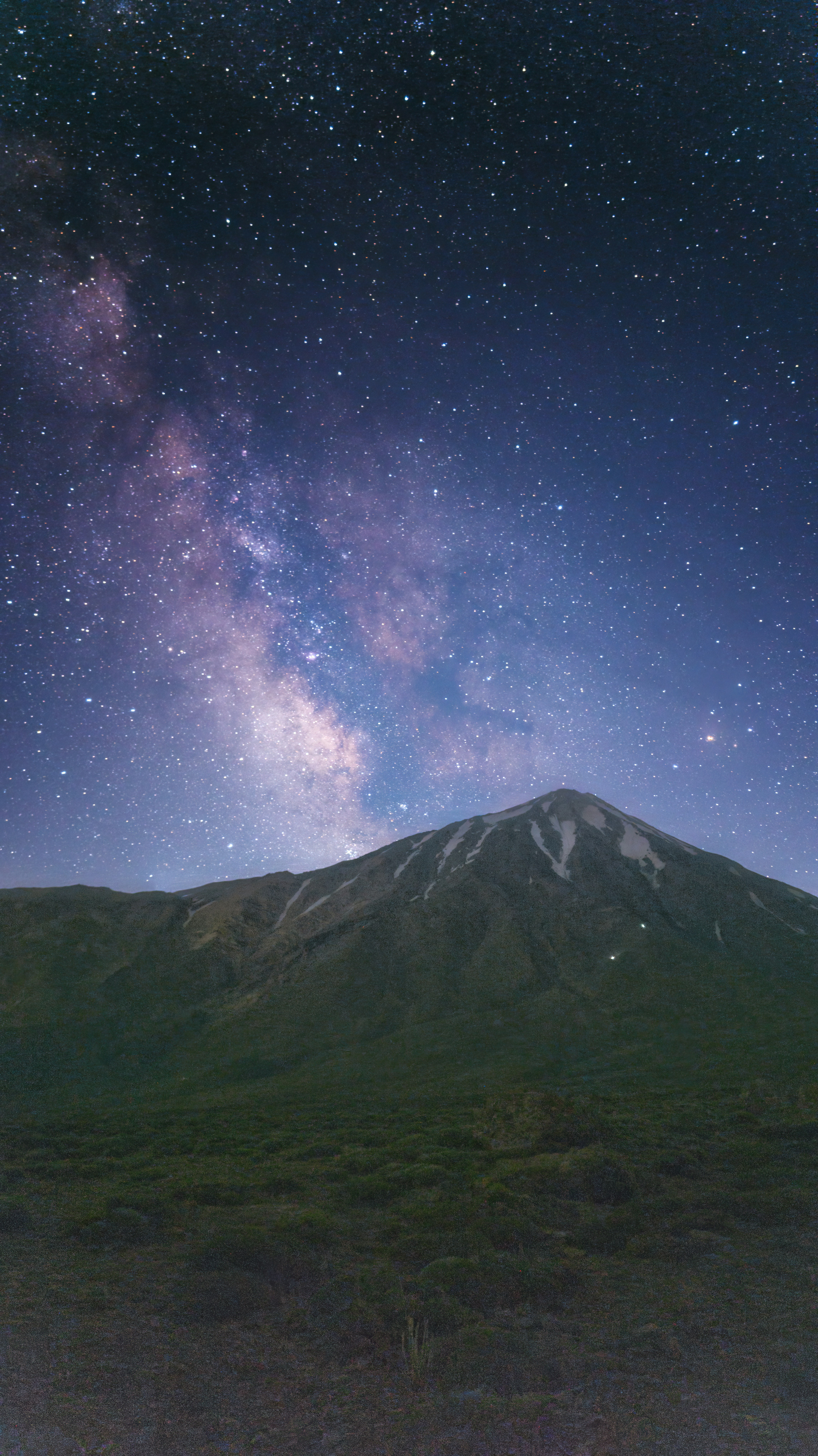 Star-filled night sky above a quiet volcano, with the Milky Way stretching across the upper atmosphere and the dark, rugged slopes below.