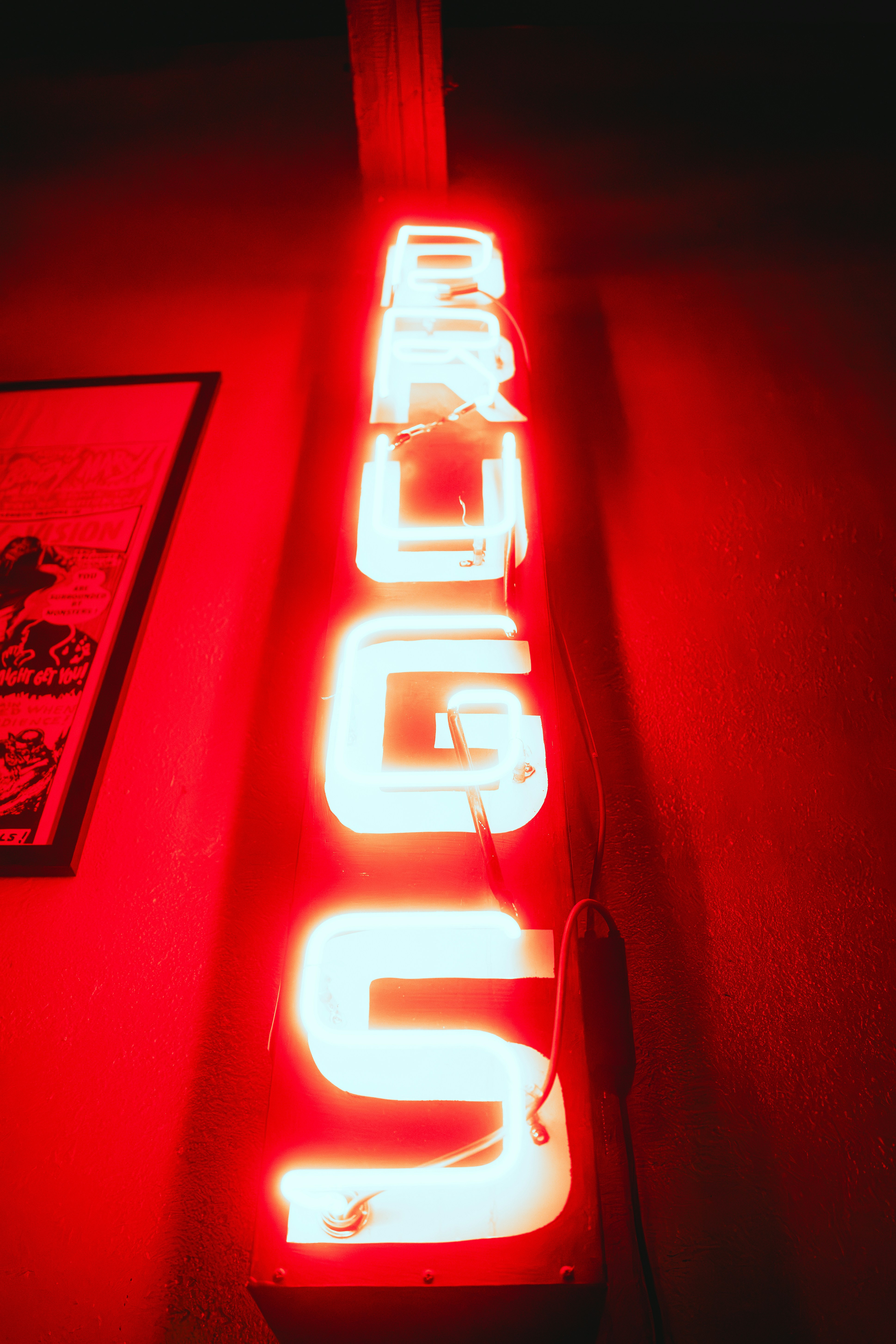 A vertical red neon sign illuminates a dim interior, casting a vivid glow on a framed poster and surrounding walls.