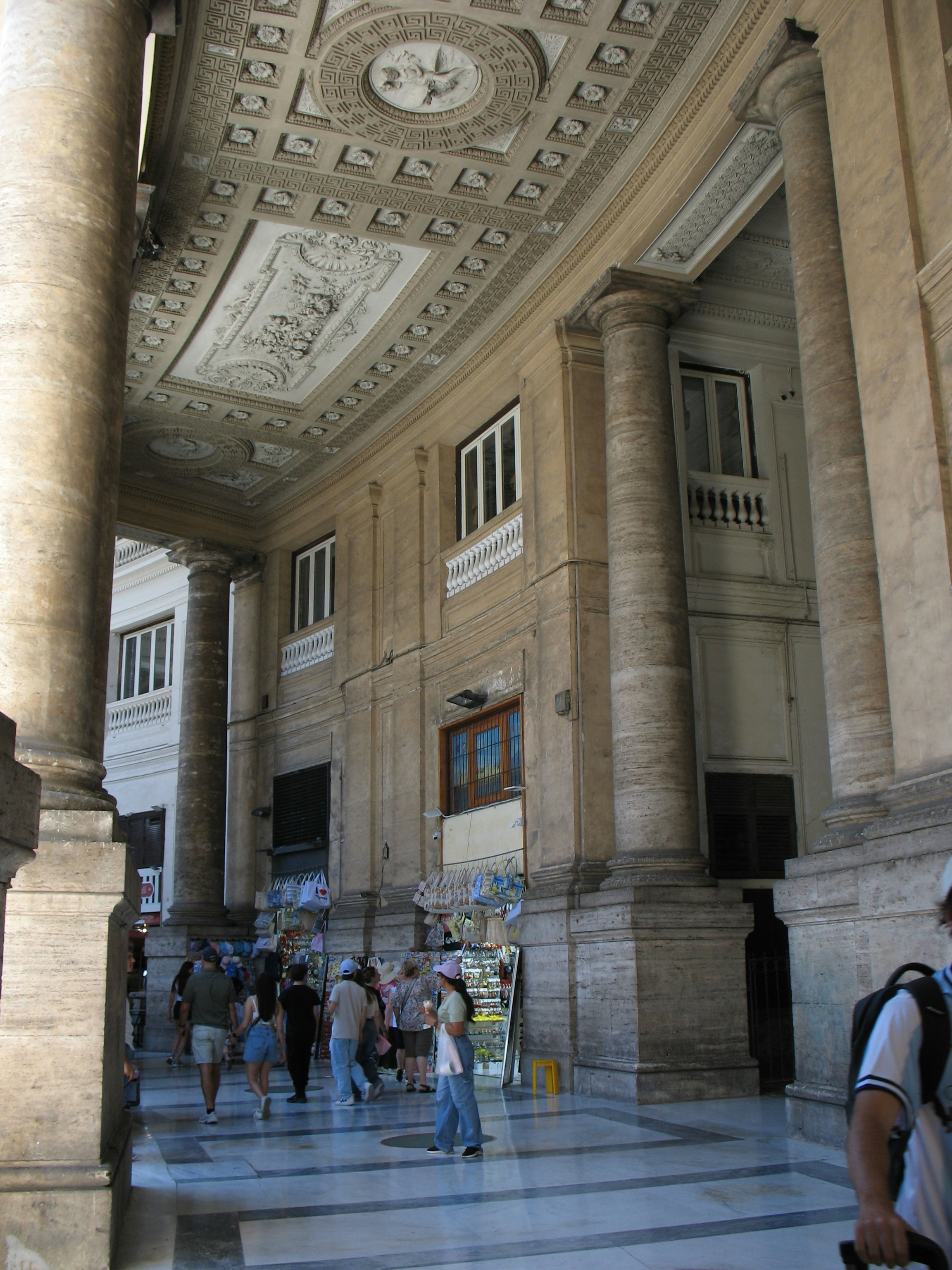 Arched colonnade with ornate ceiling relief and shoppers beneath, highlighting a grand architectural hall and street-level stalls.