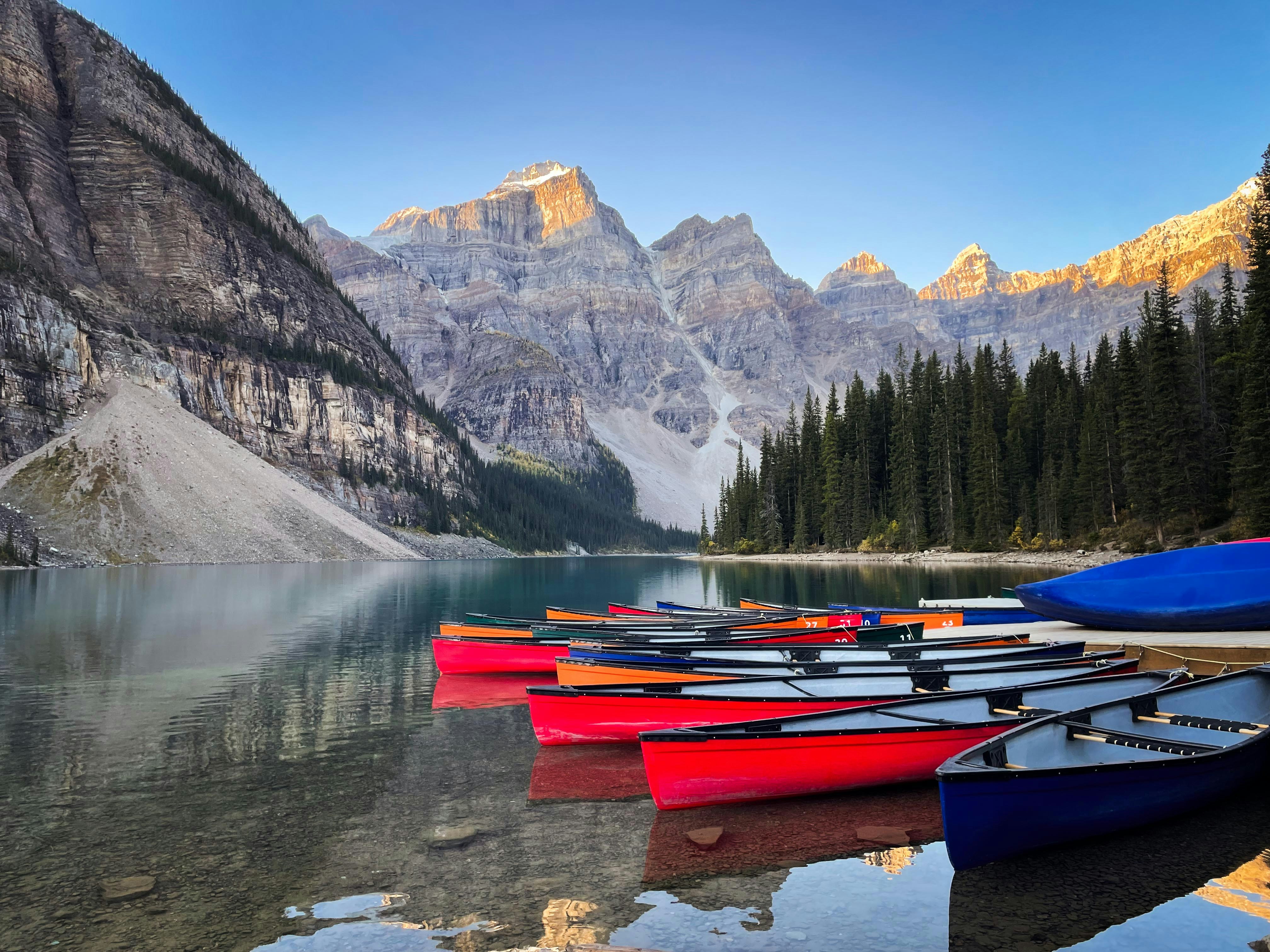 Colorful canoes line the glassy lake against a backdrop of rugged mountains and evergreen forest. A tranquil alpine scene emphasizing still water, reflections, and distant peaks.