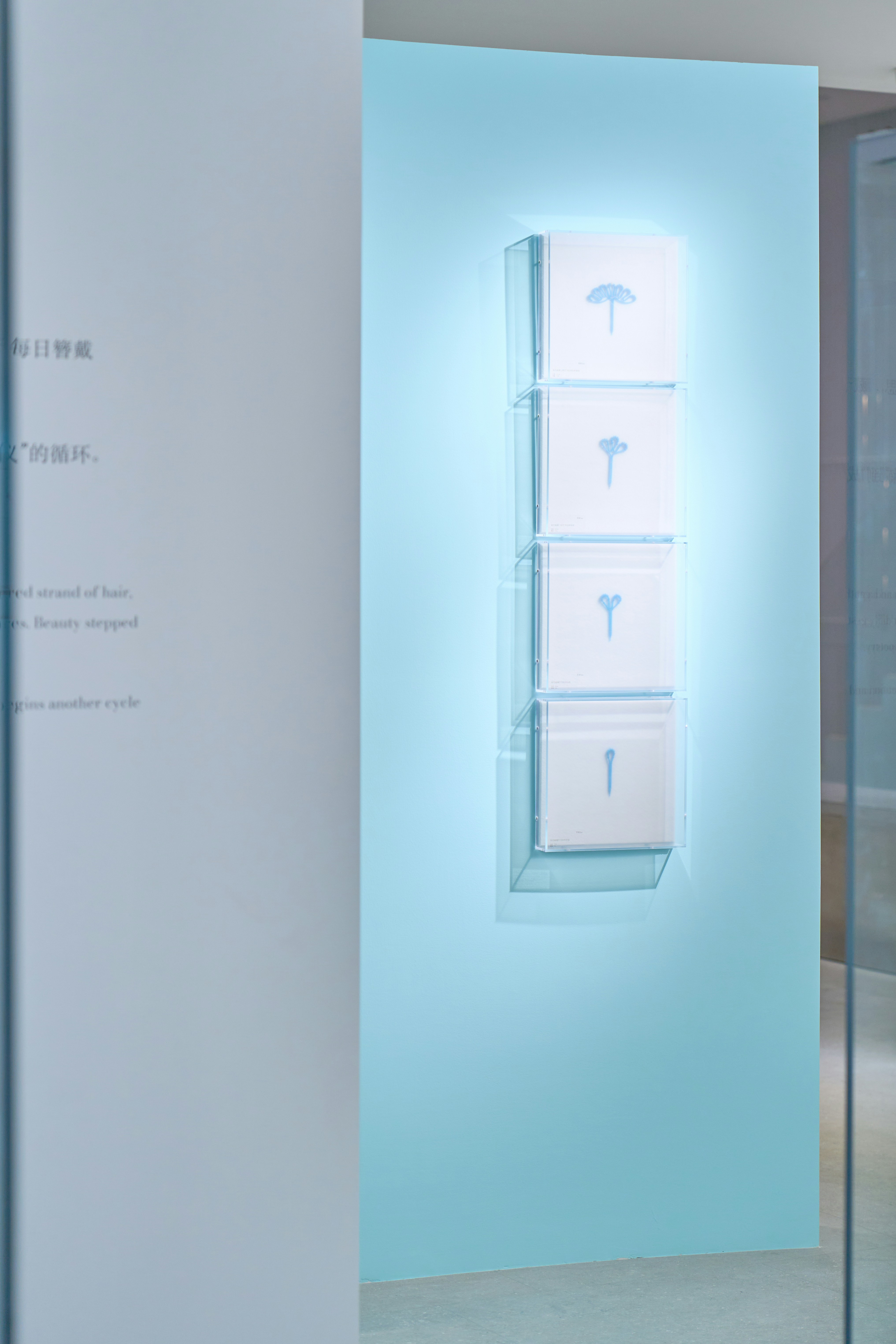 A vertical light-blue wall features a stacked, illuminated cuboid sculpture with subtle icons; the soft glow and surrounding space emphasize a minimalist gallery installation.