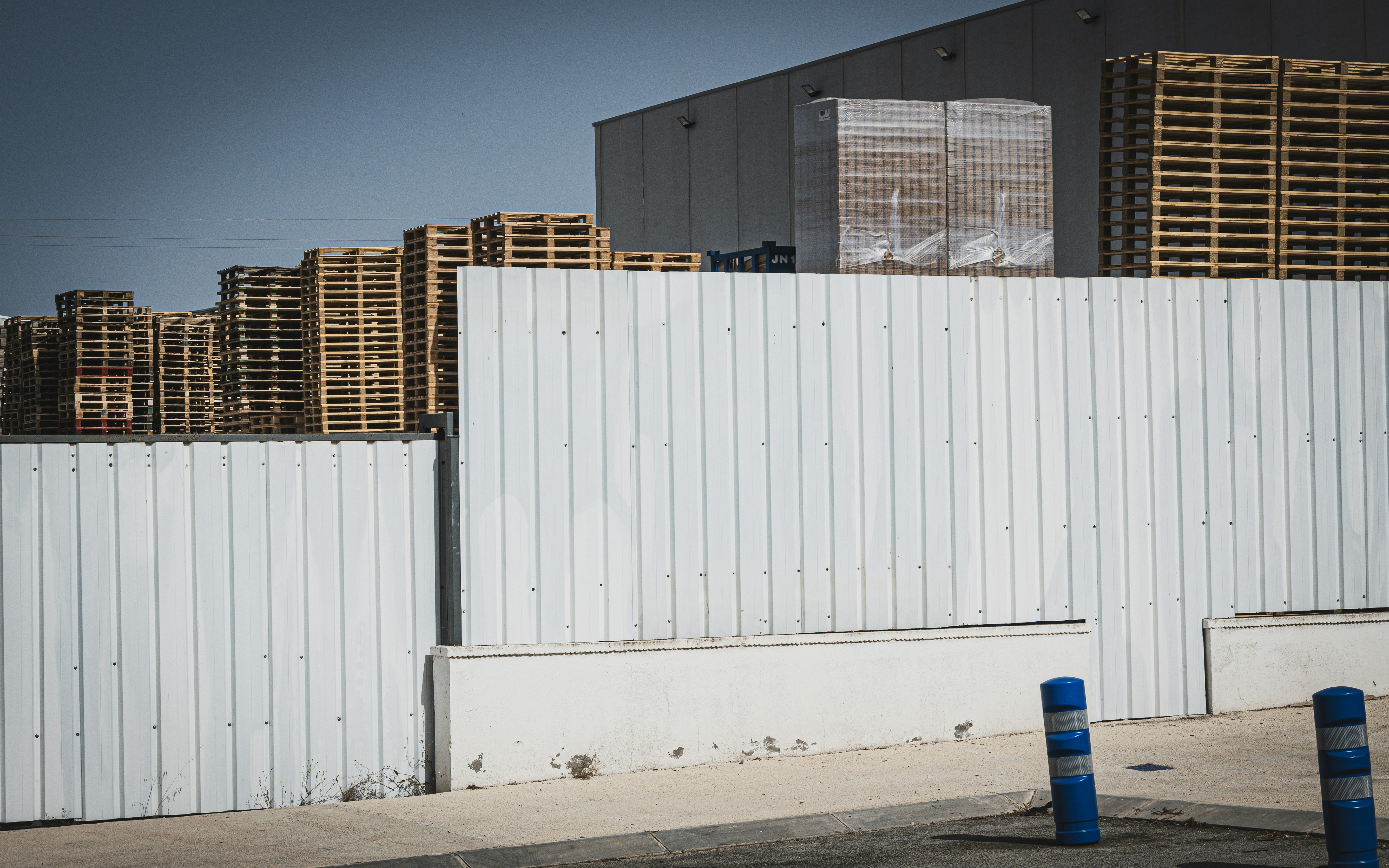 A corrugated metal wall, painted the white of institutional erasure, runs across the frame like a sentence interrupted mid-thought. Behind it, stacked wooden pallets rise in irregular towers, showing a silent side of commerce, patient and indifferent over a weekend. A grey warehouse anchors the background, its bulk softened by midday haze, while two blue bollards stand sentinel on the empty forecourt below. The scene recalls de Chirico's plazas: geometries of waiting, drained of human occasion.