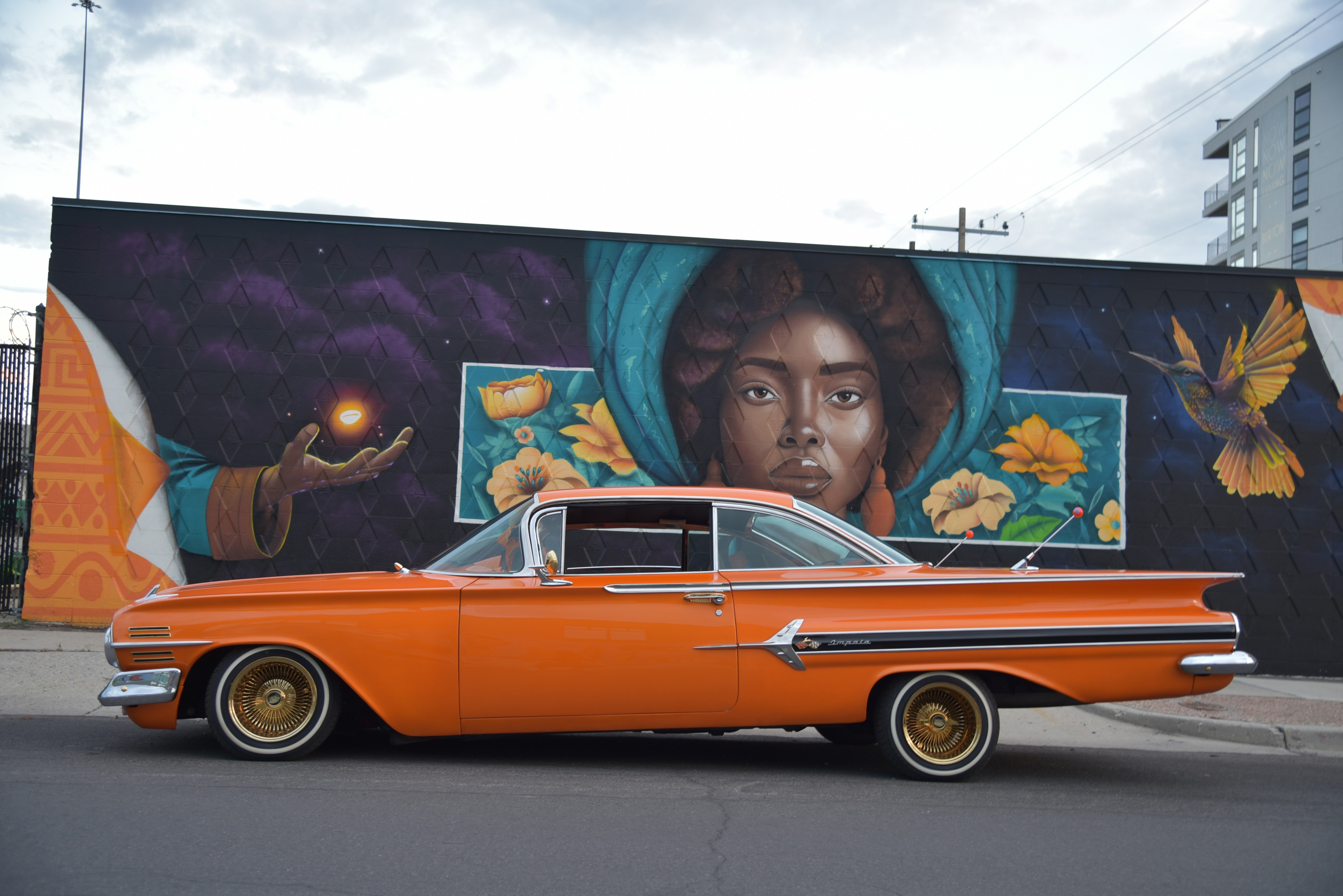 Orange vintage car parked before a vivid mural featuring a woman's face and tropical birds; street scene emphasizes color and retro vibe.