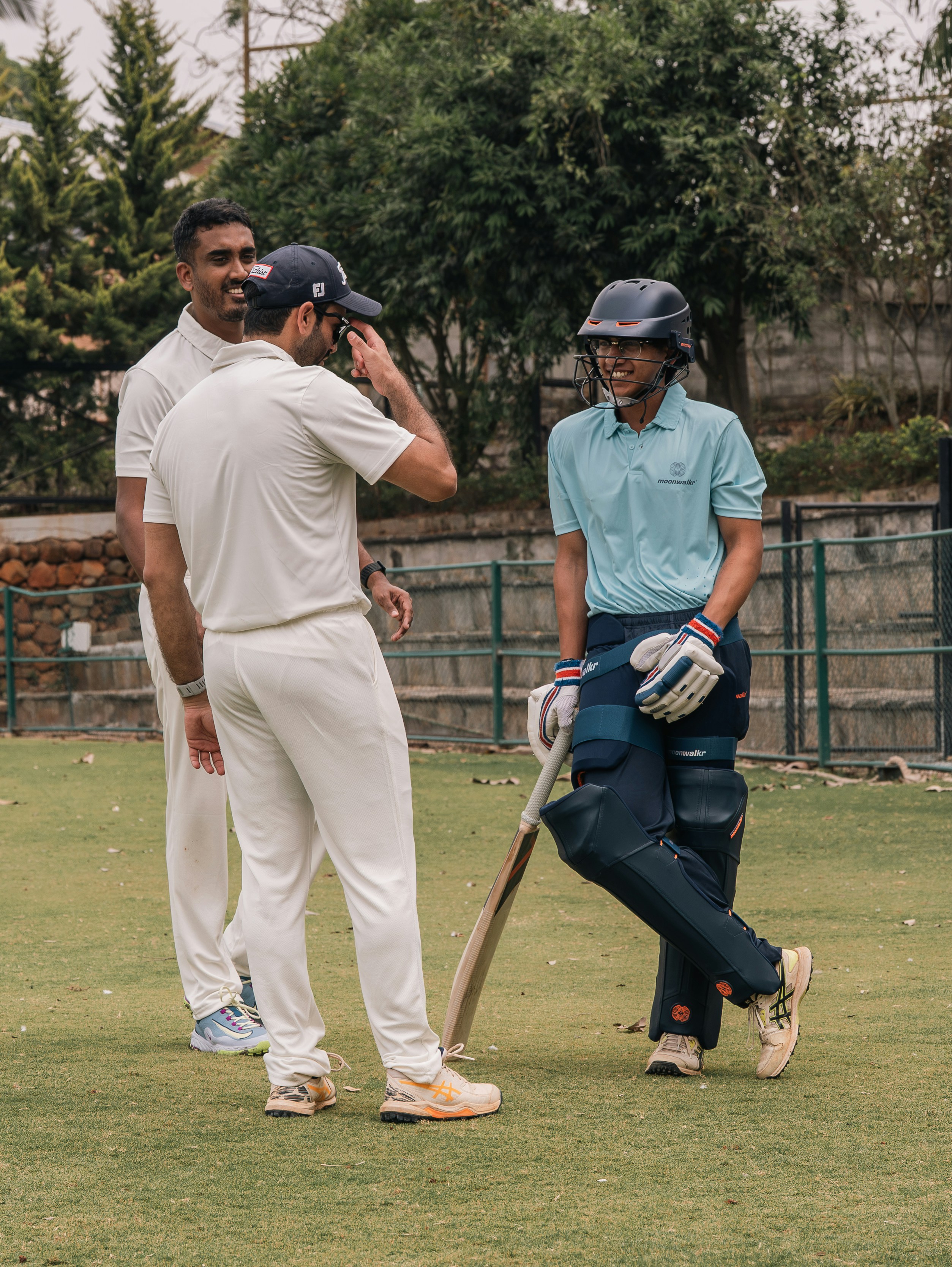 Three cricketers stand on a grassy field, one wearing a helmet and leg guards while another adjusts his cap, engaging in conversation near the batsman in a relaxed moment during practice. This is a candid scene captured outdoors.