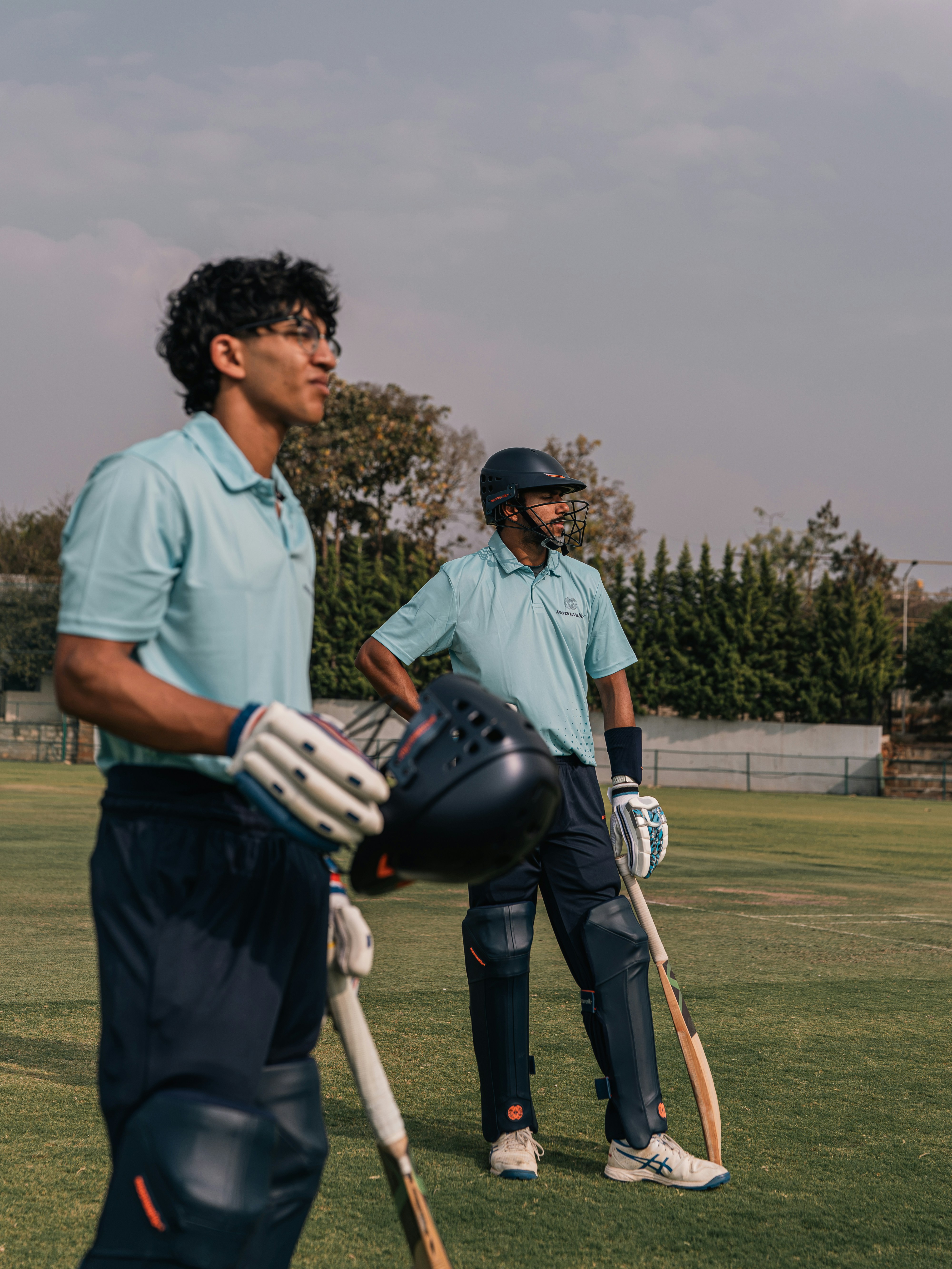 Two cricket players on a sunlit field pause for a moment during practice