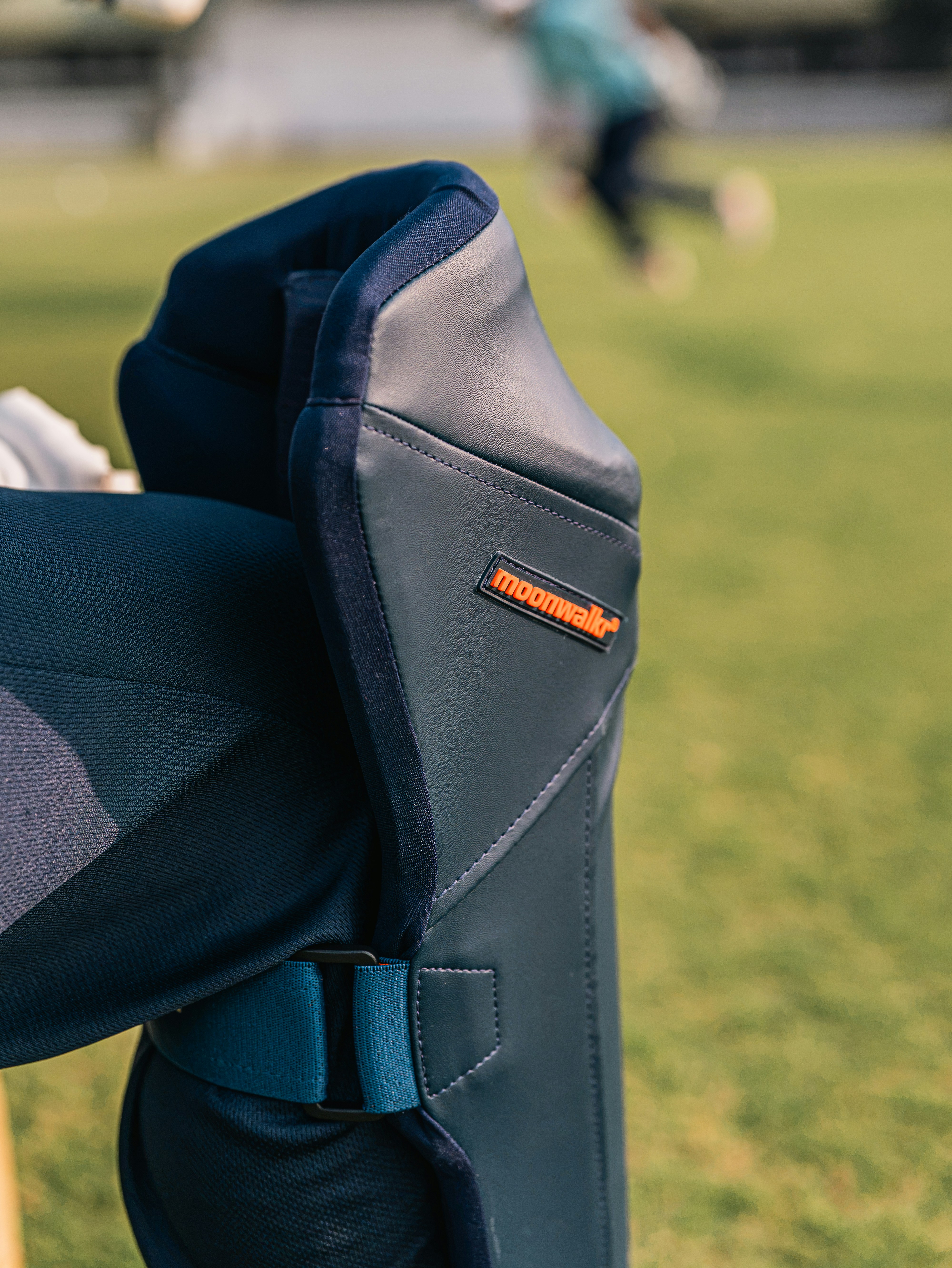 Close-up of a dark blue padded vest with a small orange logo, resting on a bench or hook in an outdoor field; background shows blurred figures and green grass, indicating a sports practice scene. This is a fabric/gear detail shot.