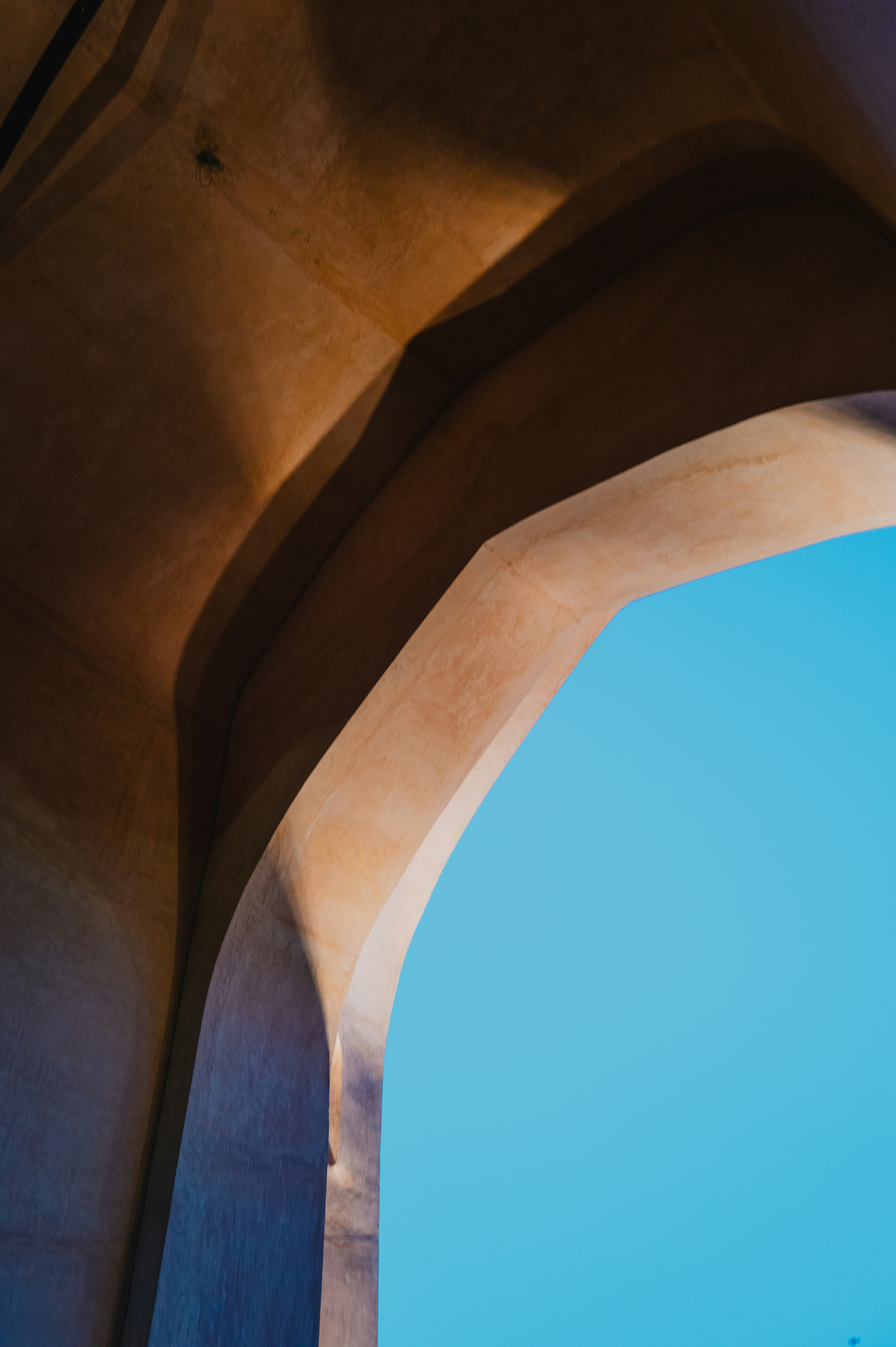 Warm stone curves frame a crisp blue sky through a large arch, emphasizing geometric architecture and shadow lines.
