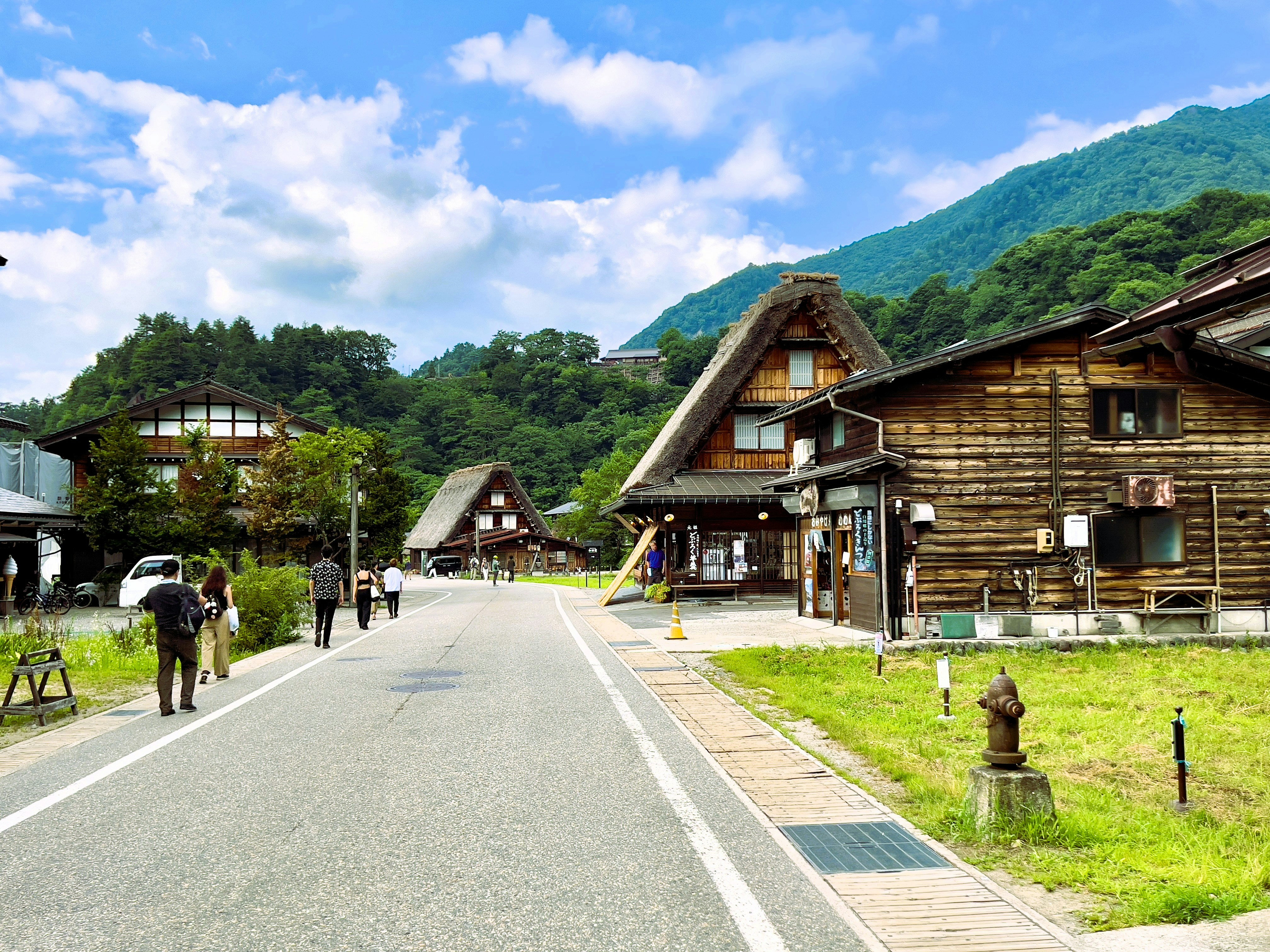 A quiet village street with traditional wooden houses lined along a mountain backdrop, pedestrians strolling and a bright, clear sky above.