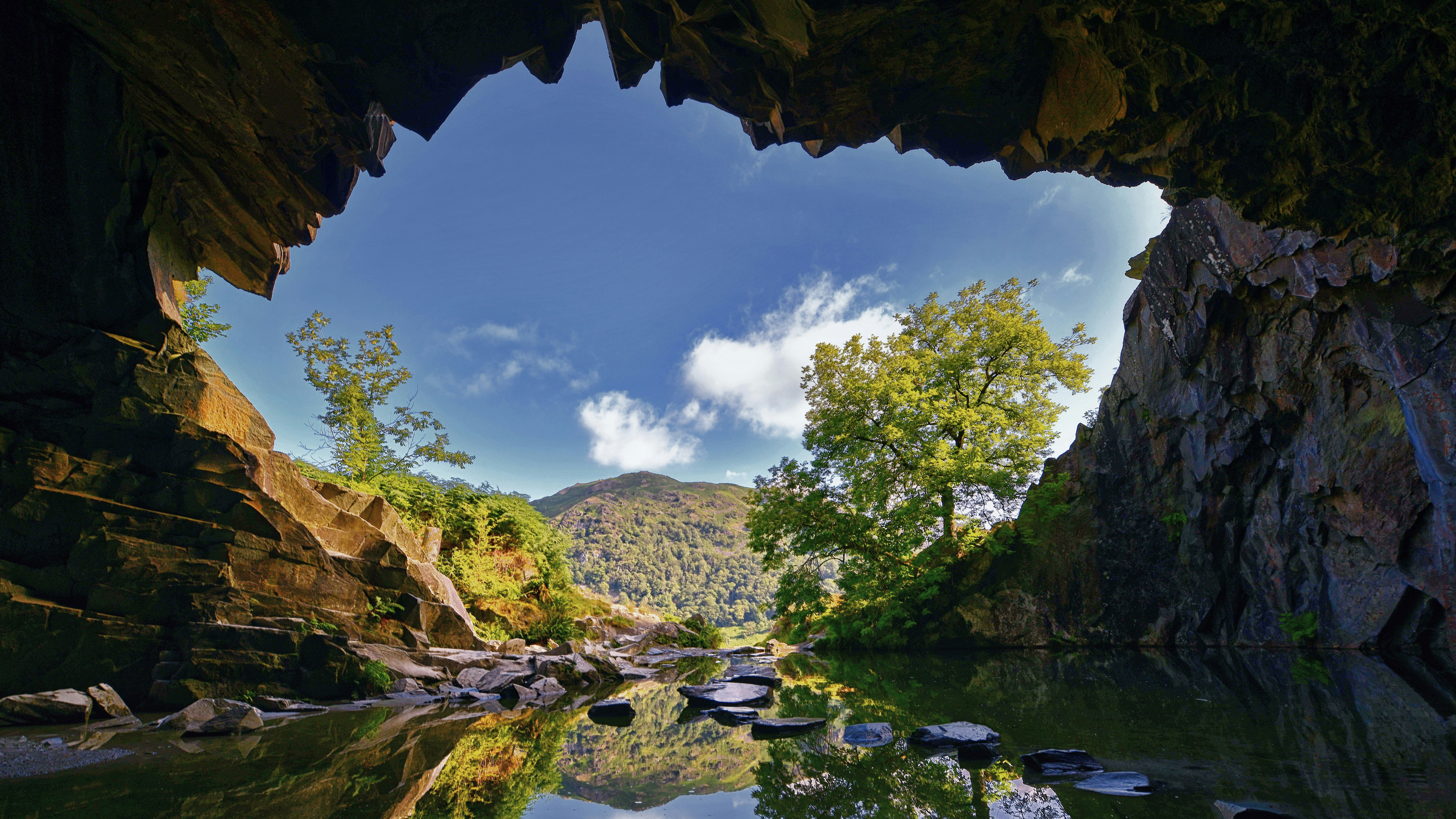 Looking out from the entrance of Rydal Cave towards the stepping stones and surrounding fells in England's Lake District National Park. The natural rock arch frames a peaceful landscape shaped by centuries of quarrying and the timeless beauty of Cumbria.