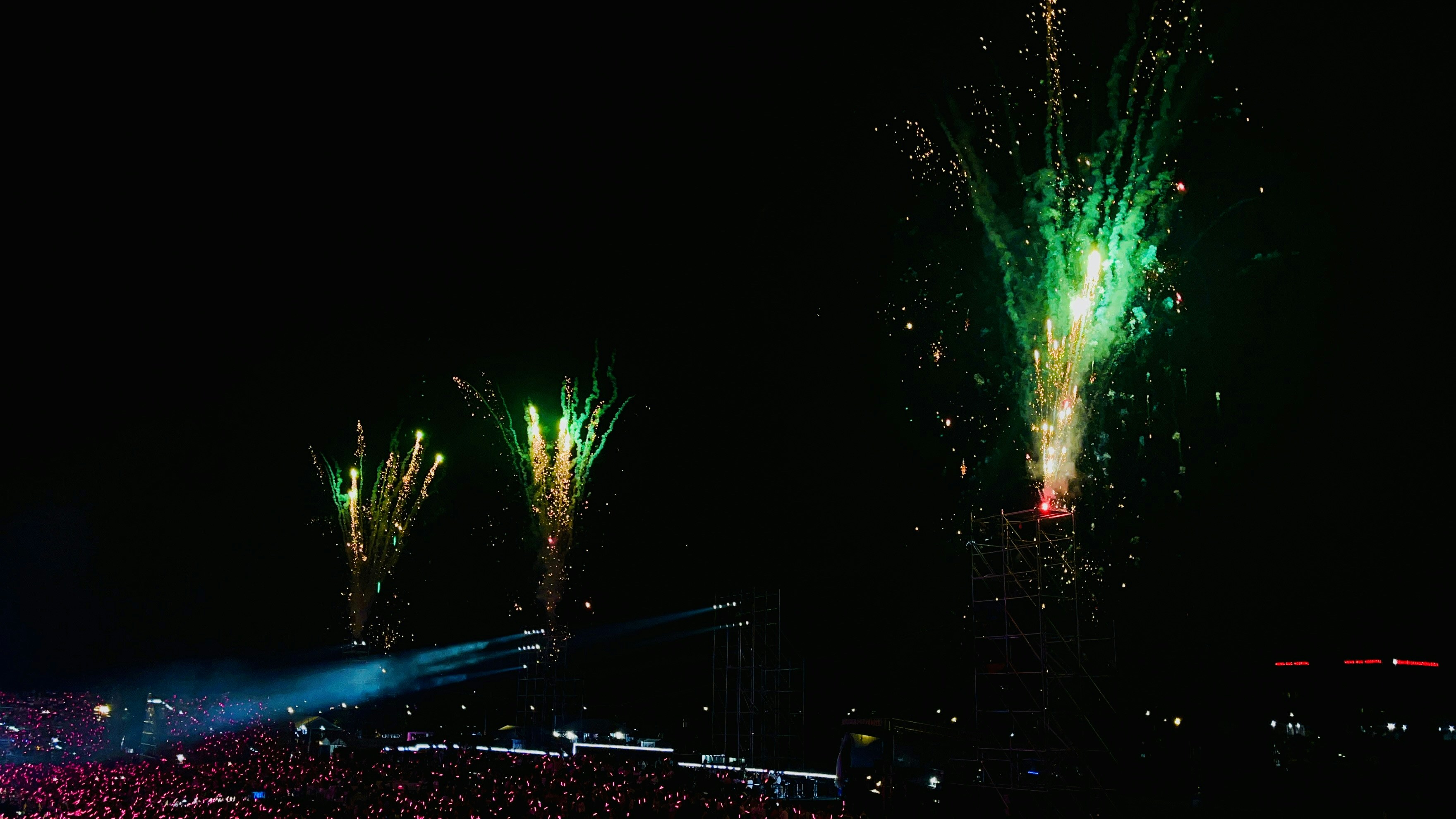 Colorful green fireworks explode above a dark riverfront with silhouettes and faint structures, creating a dramatic nighttime scene.