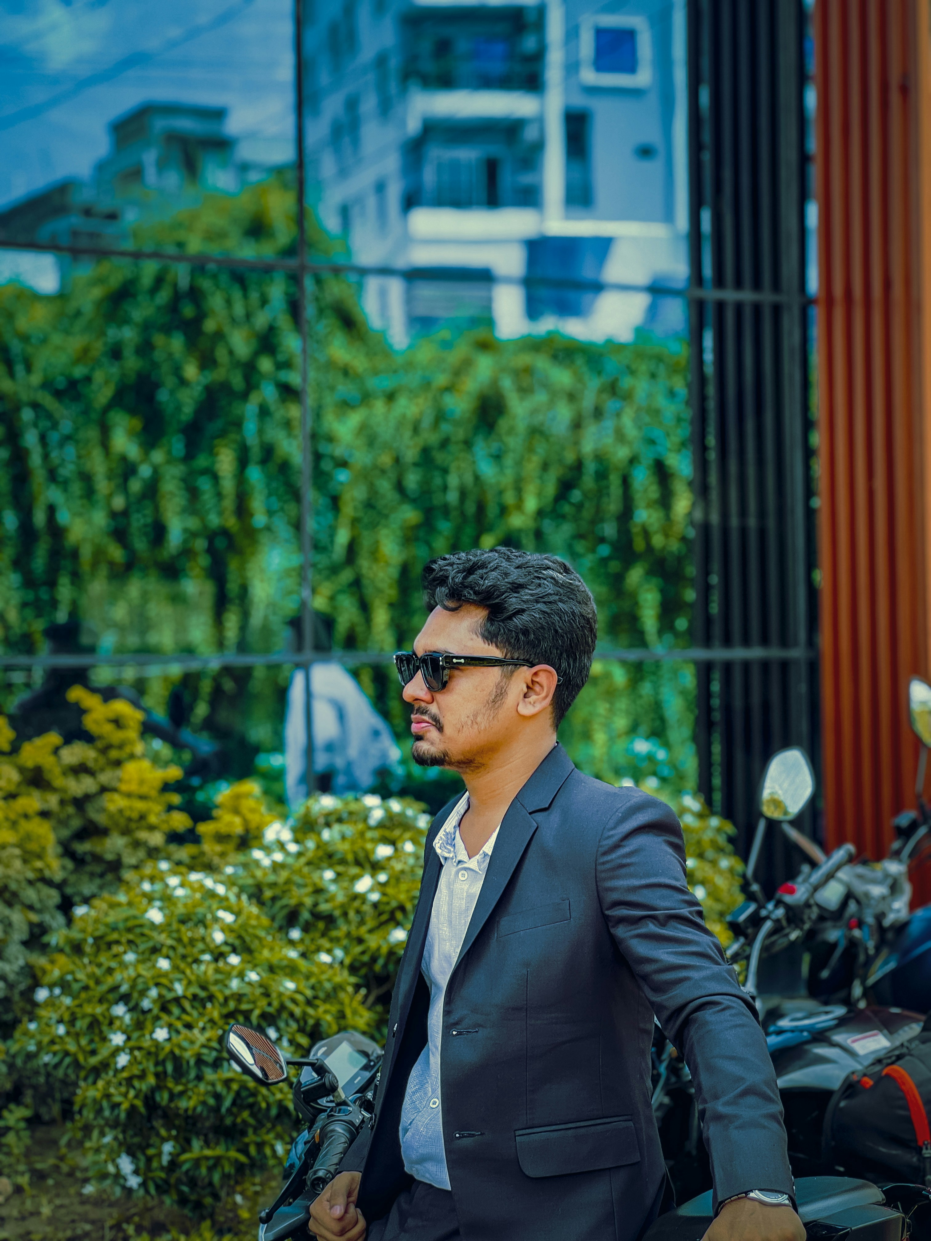Man in a dark suit and white shirt stands beside motorcycles in front of a glass-fronted building and greenery.