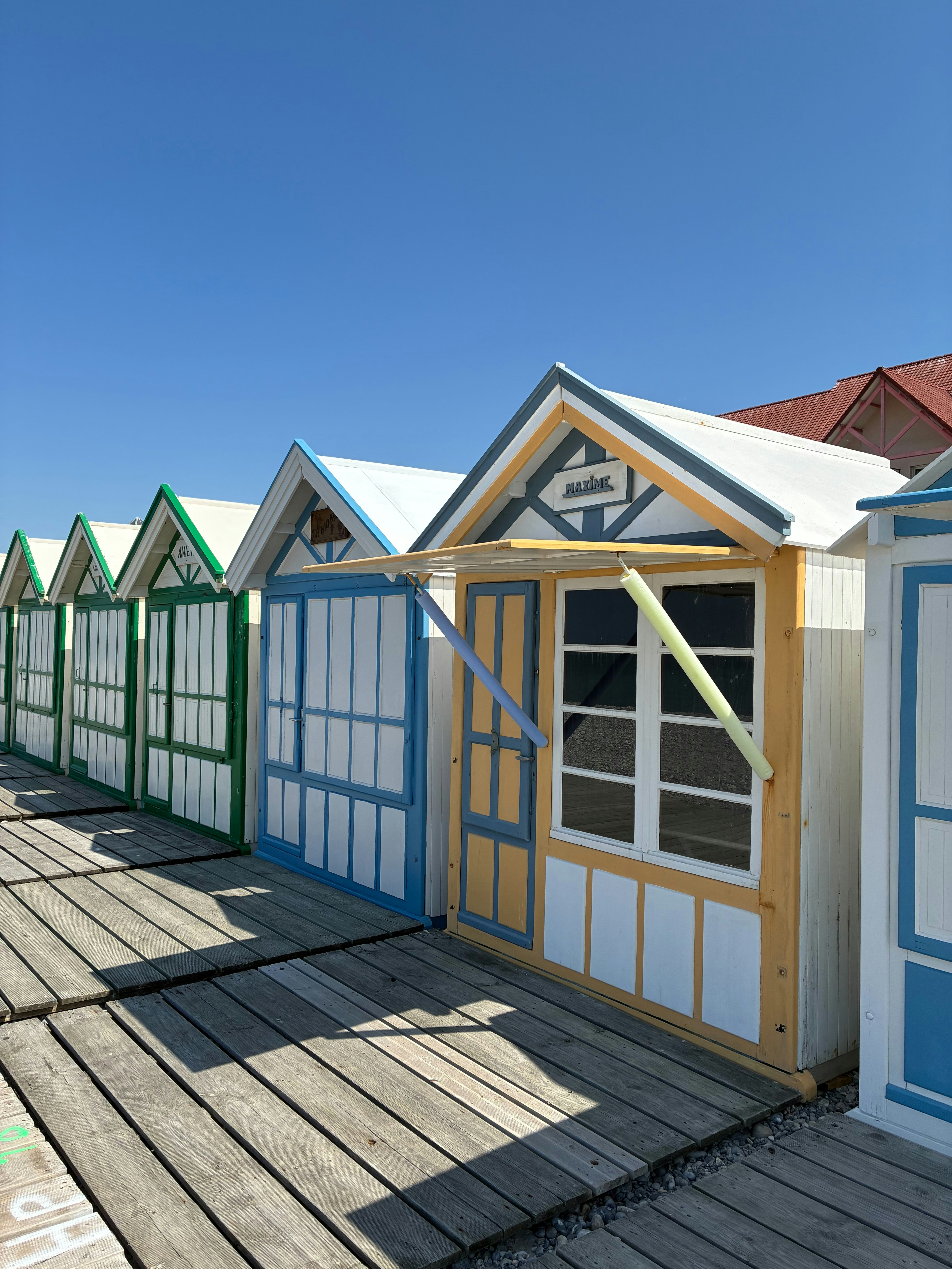 Colorful row of small beach huts with pastel blue, green, and yellow trim along a wooden boardwalk; an orderly coastal display beneath a bright sky.