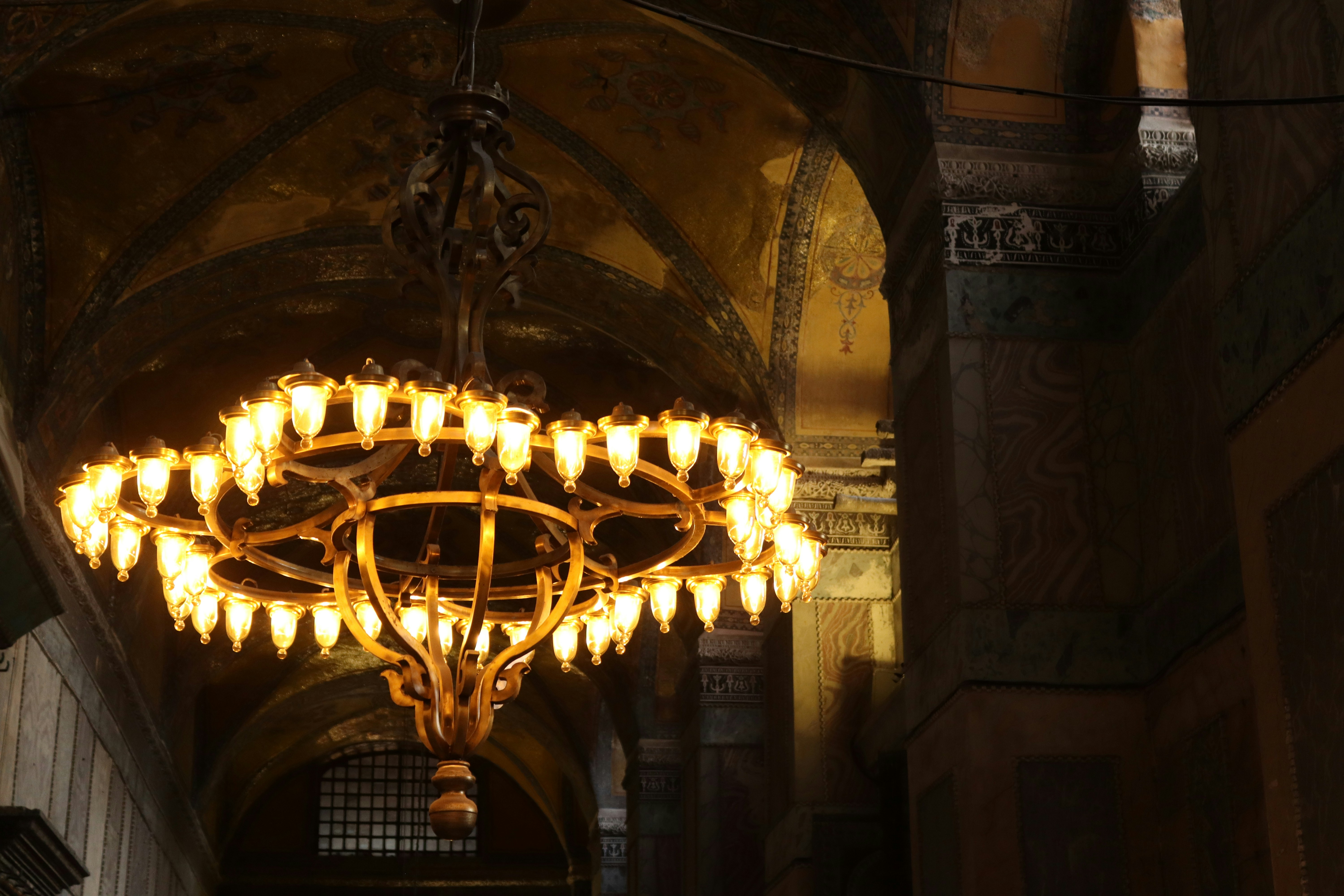 Intricately lit chandelier hangs over a dark, vaulted interior, highlighting ornate architecture and warm glow.