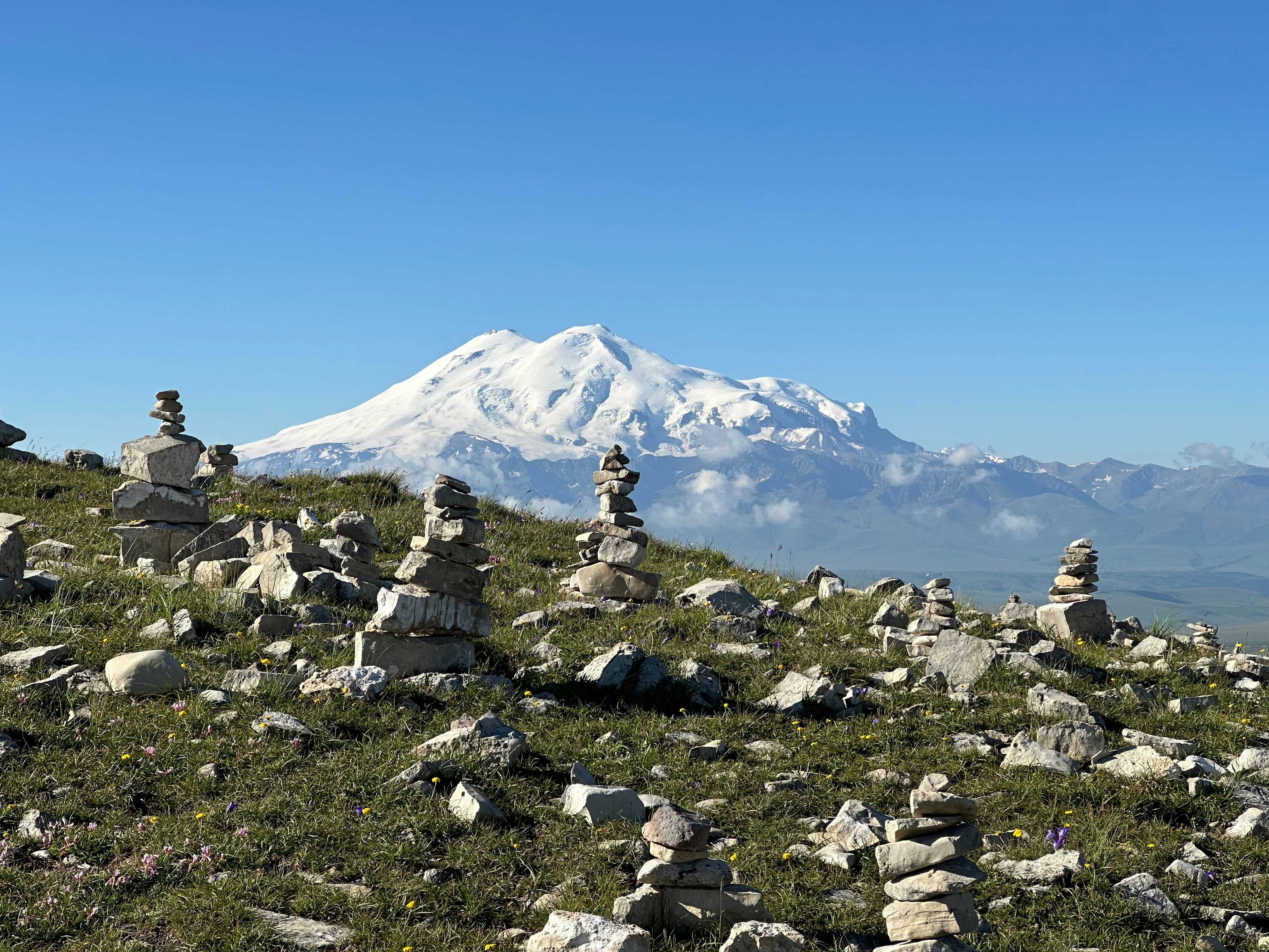 Row of stone cairns dot a grassy alpine plateau with a snow-capped peak in the distance under a bright blue sky.