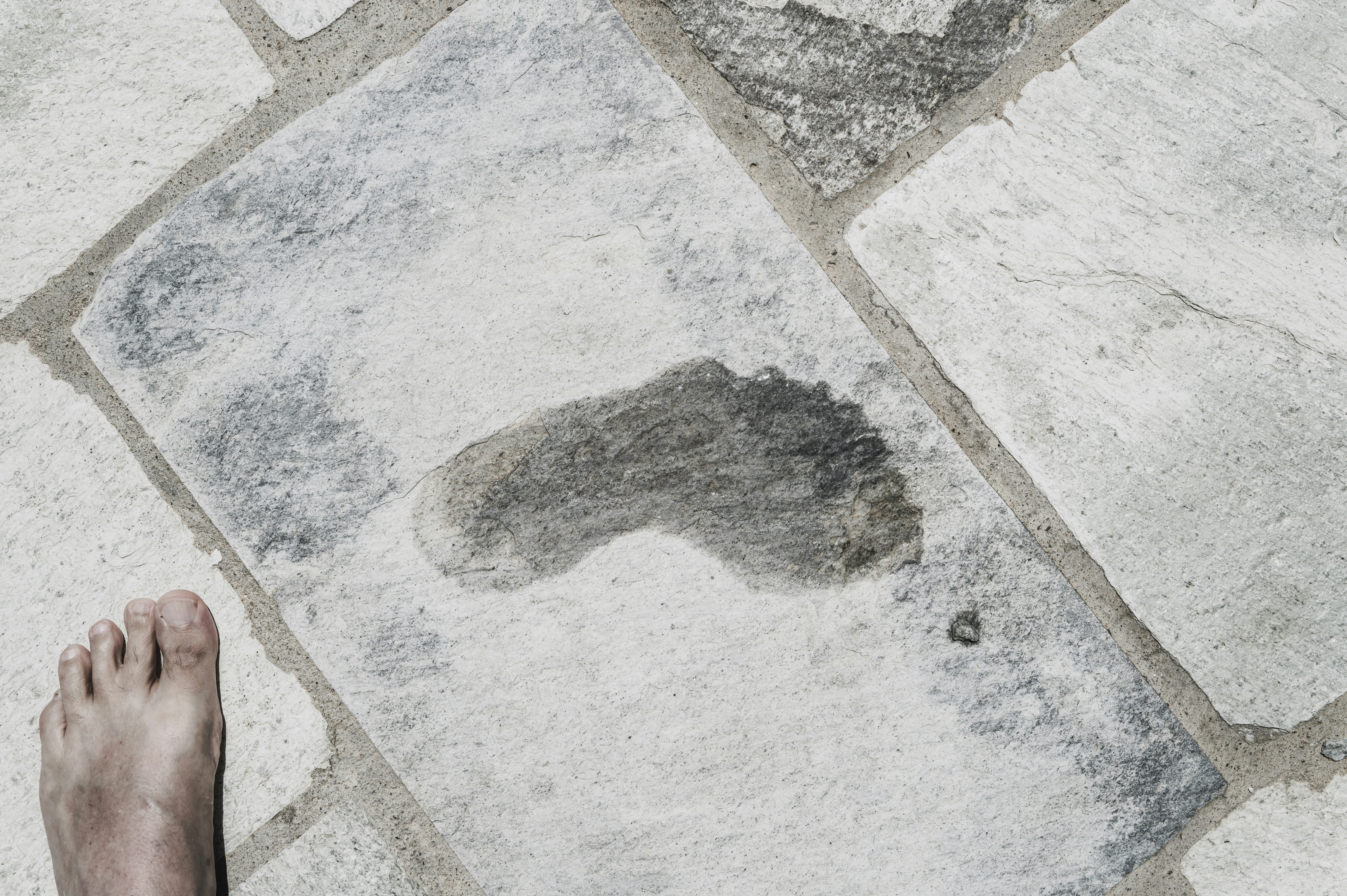 A close-up view of a weathered stone patio with a single bare foot entering from the lower-left, sunlight softening the textured surface.