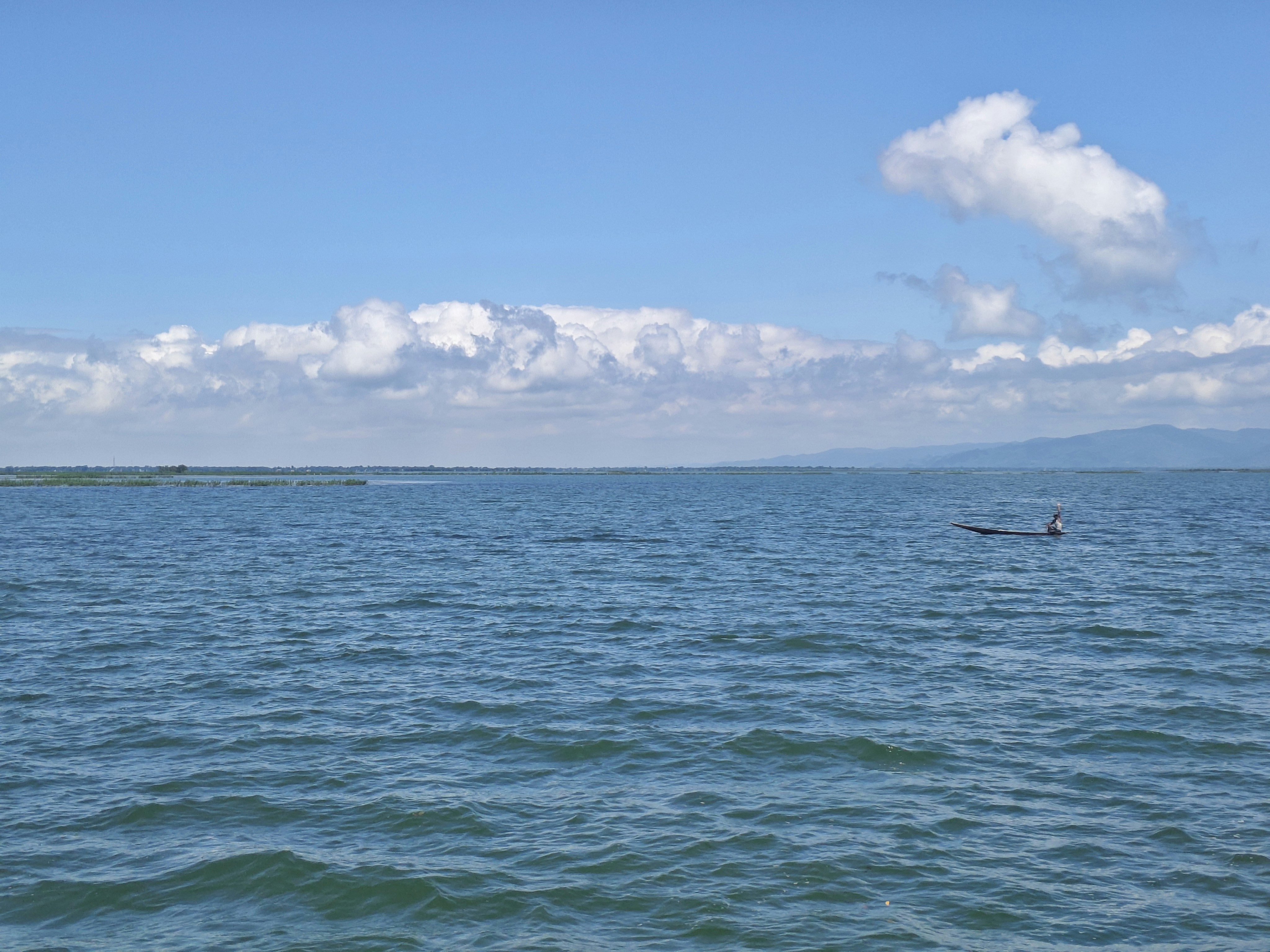 A solitary wooden boat glides across endless blue water beneath towering white clouds, creating a peaceful moment of simplicity.