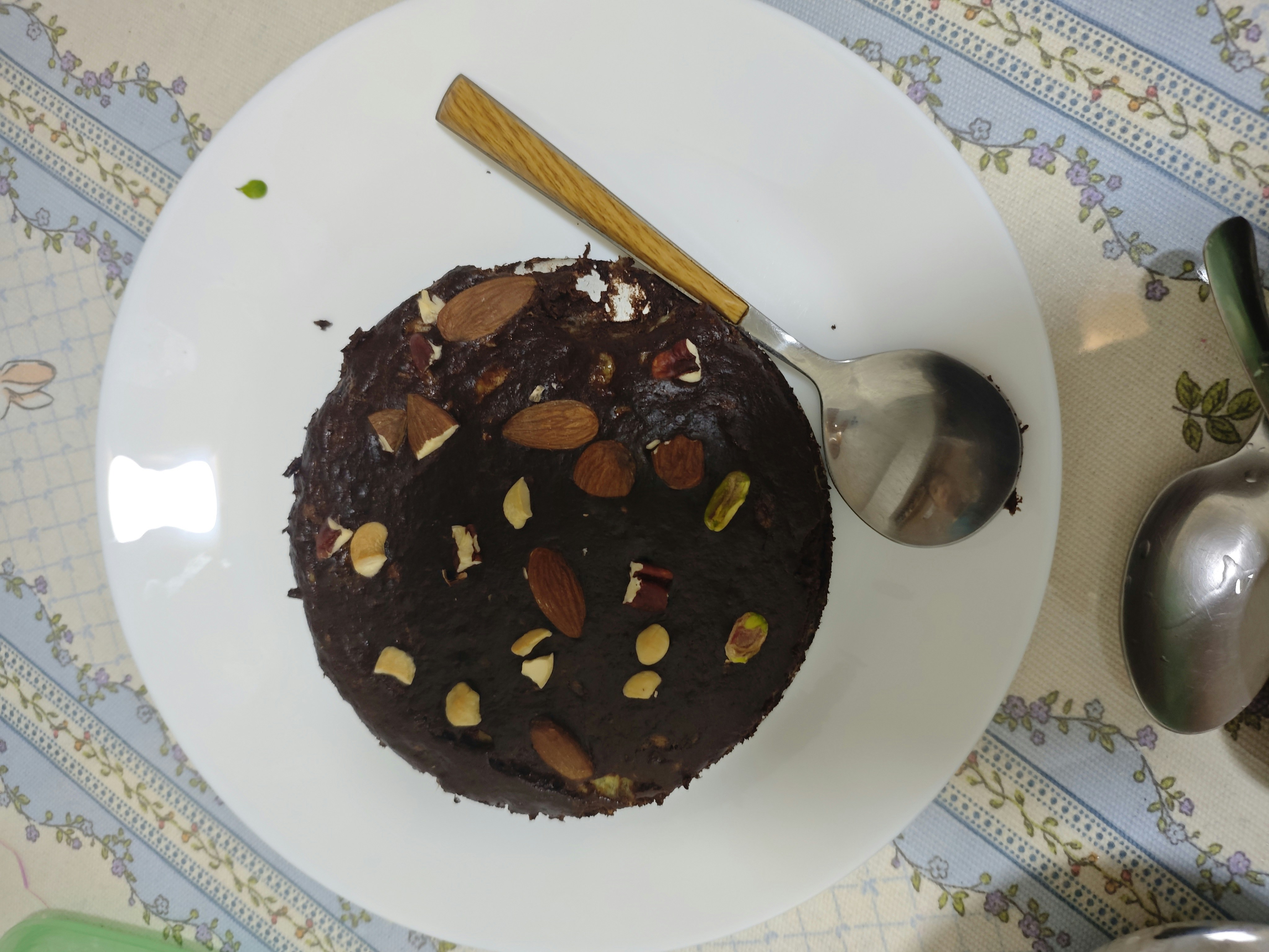 A chocolate cake topped with almond slivers sits on a white plate, with a spoon nearby on a patterned tablecloth. The scene emphasizes the dessert as the focal point.