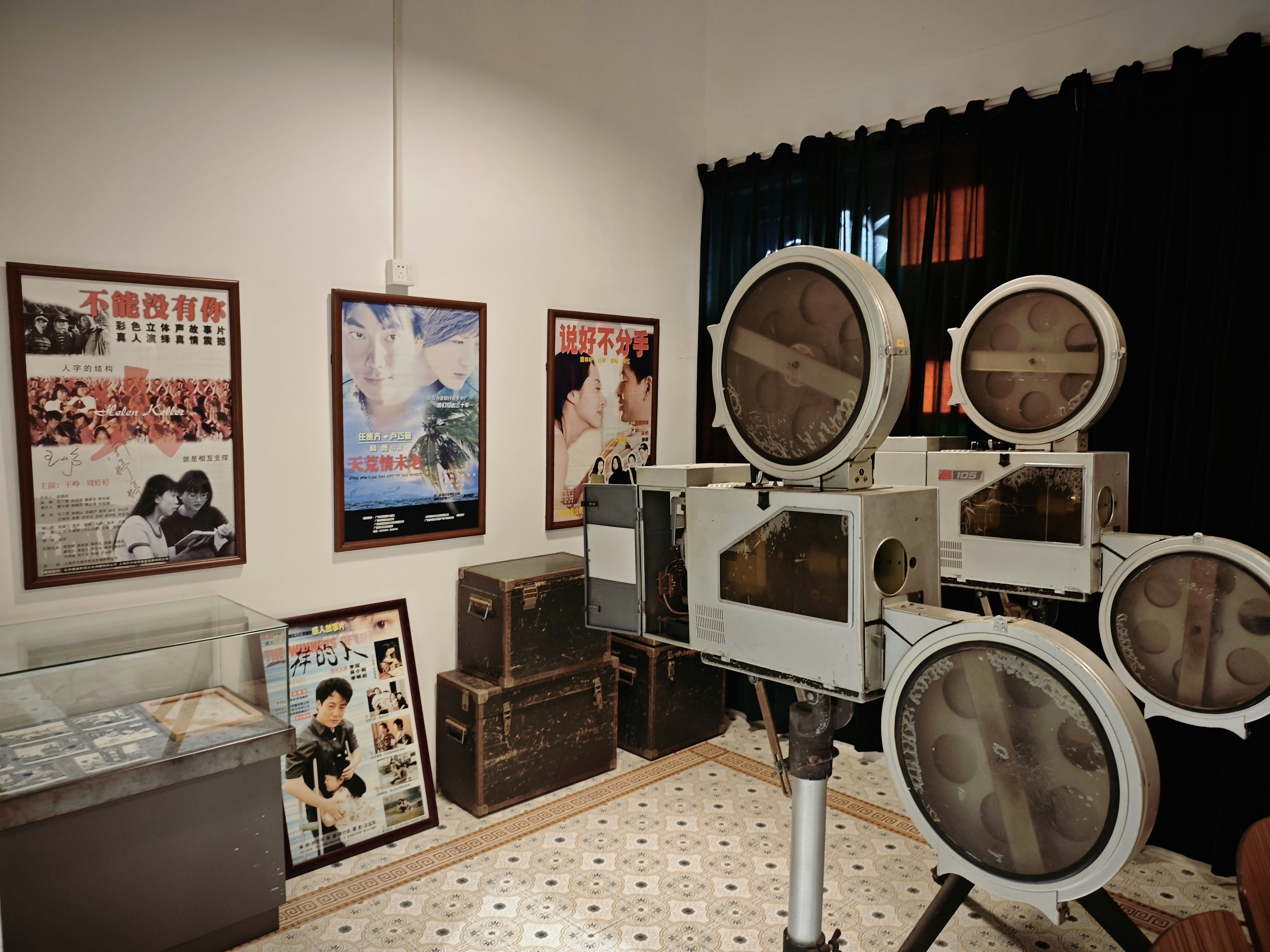 A retro film processing room featuring large circular viewing lamps and vintage posters on the wall, arranged around a glass display and stacked cases. This is a staged exhibit or studio setup with aged equipment.