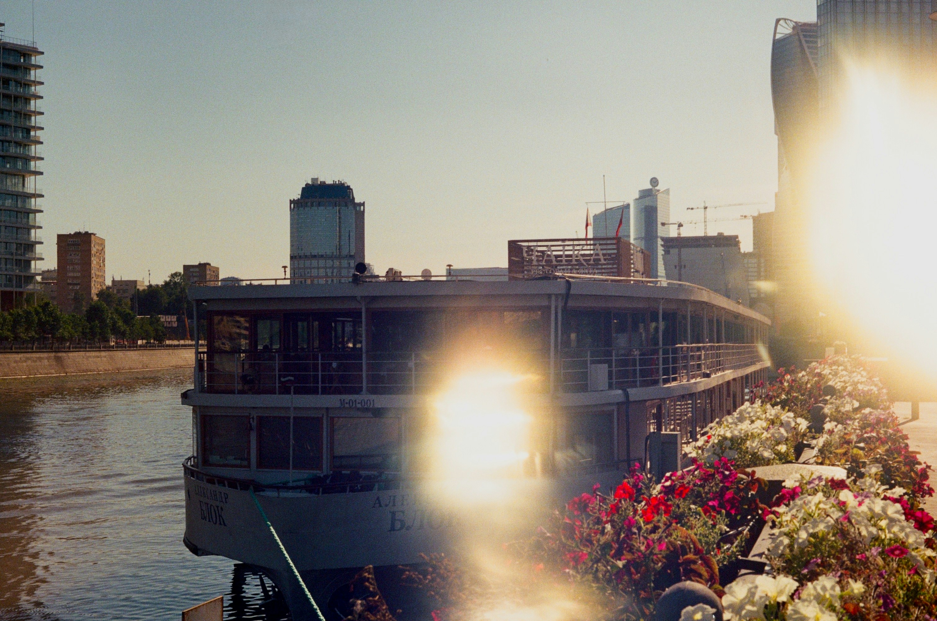 Sunlit riverfront with a multi-deck boat beside a flower-filled railing, river and city skyline in the background.