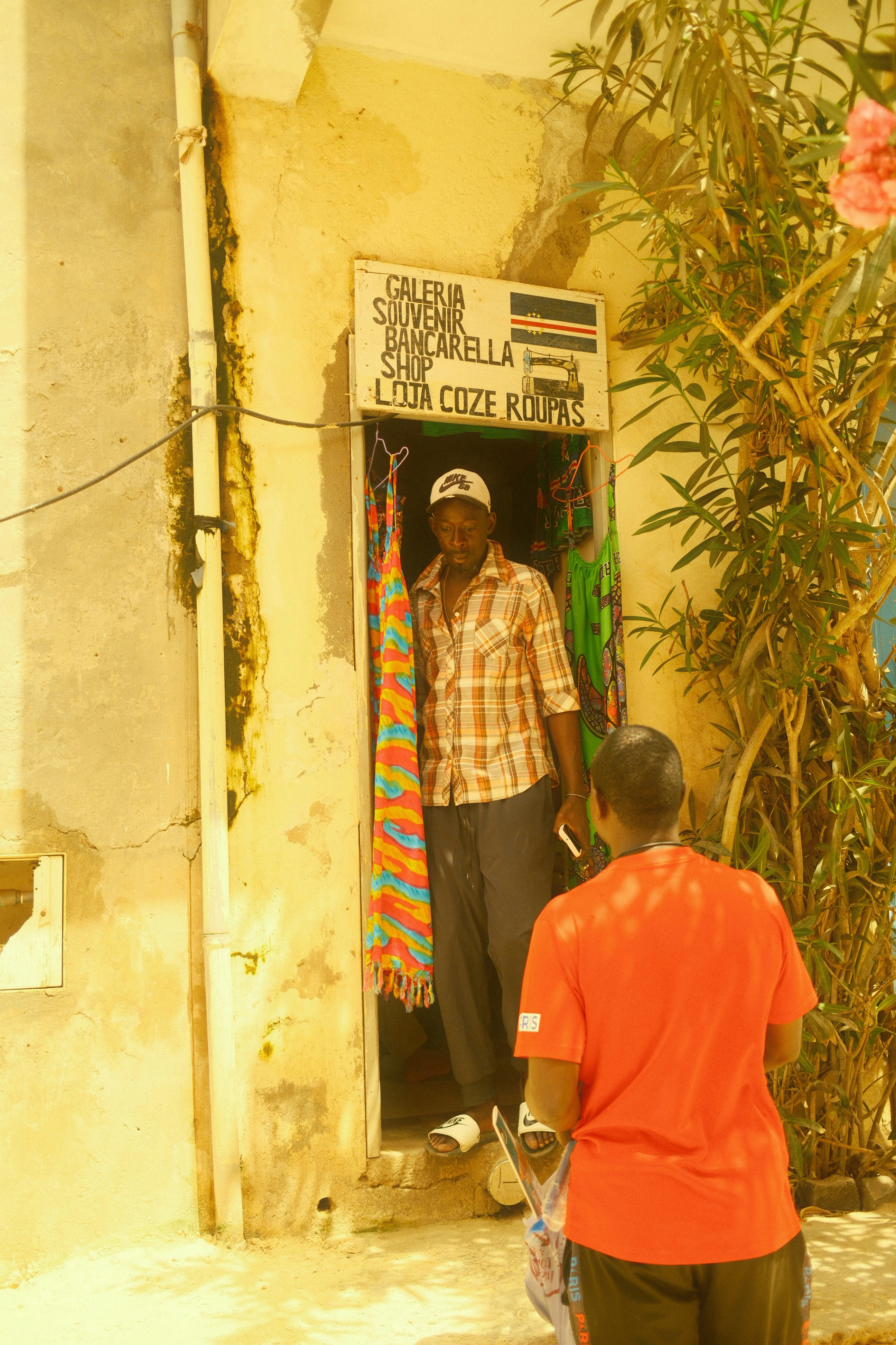 Market Entrance: A vendor in plaid greets a passerby at a sunlit doorway
